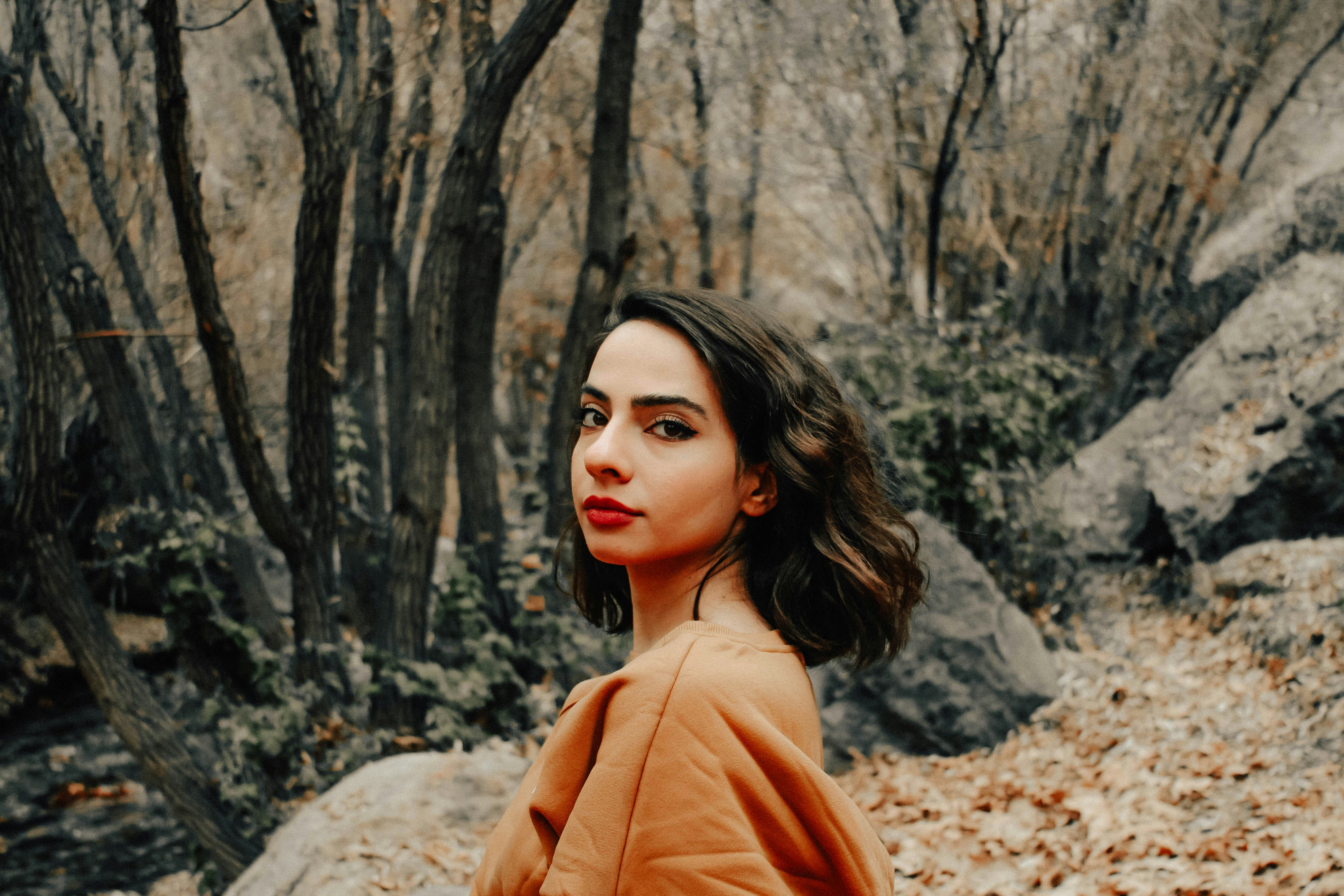 Woman in an orange coat standing amidst bare trees and fallen leaves in a forest.