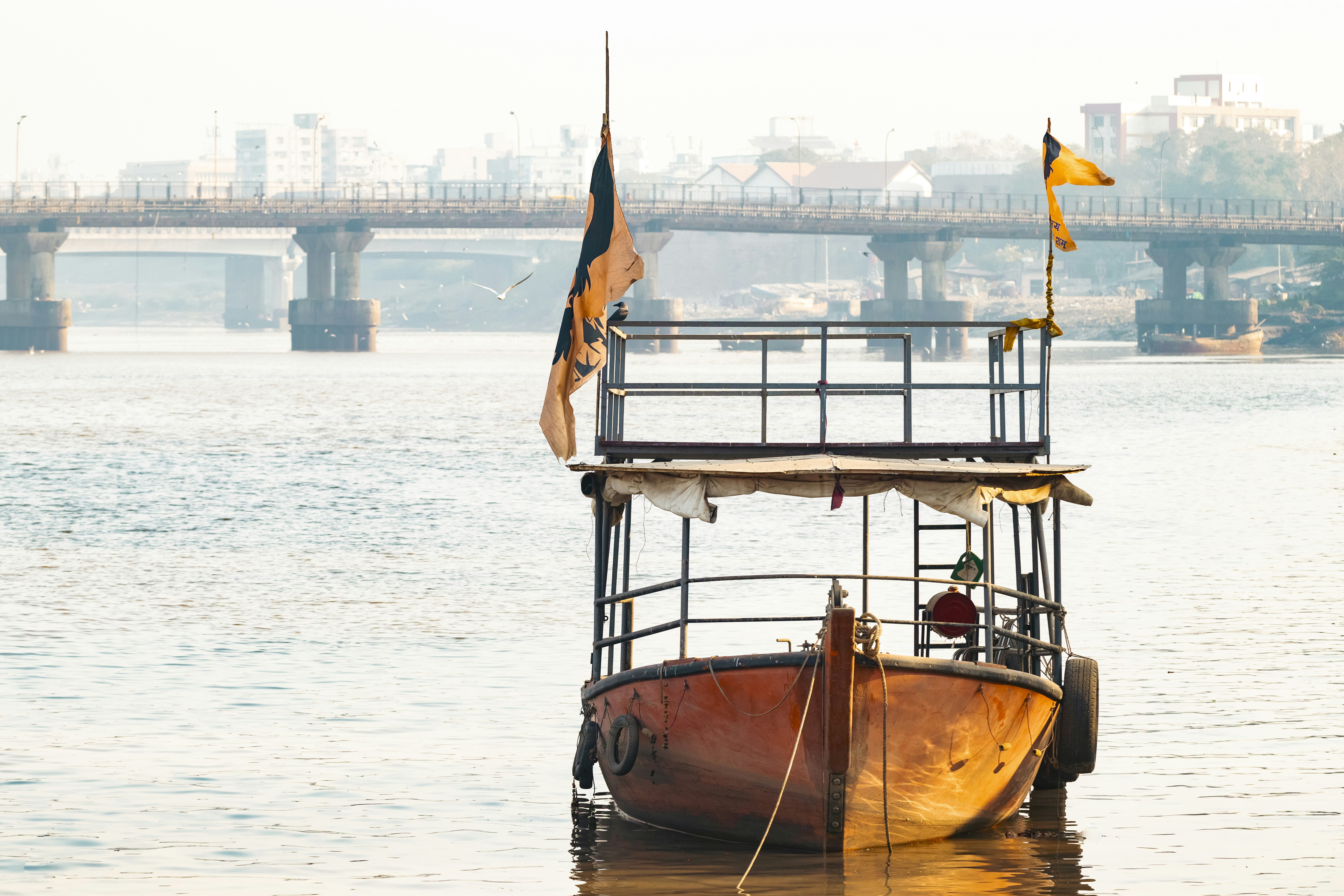 A wooden boat adorned with flags floats serenely on the Tapi River, with a bridge and cityscape in the background.