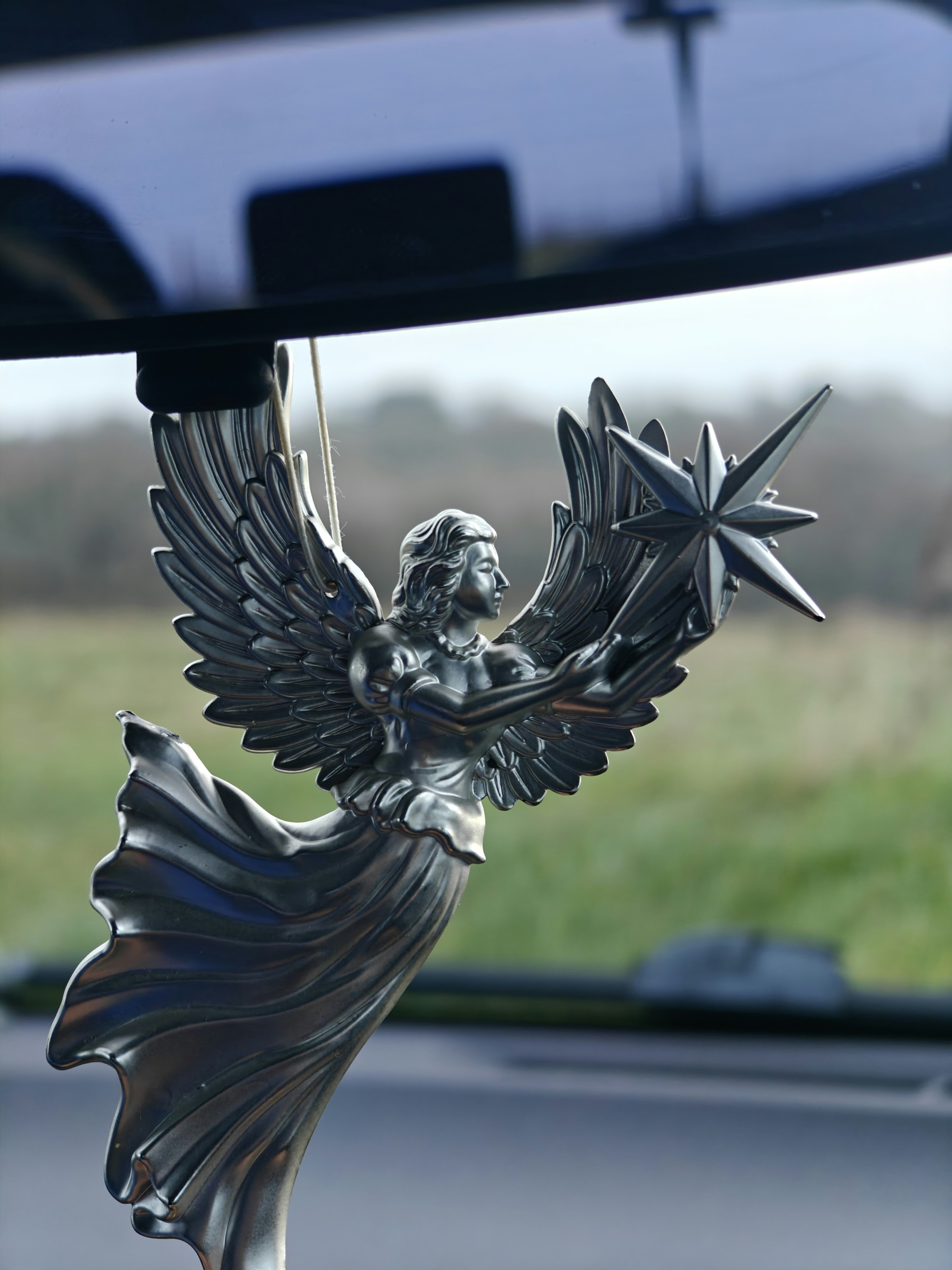 A metallic winged angel ornament hangs from a car’s rearview mirror. Its outstretched arm holds a star against a softly blurred countryside backdrop.
