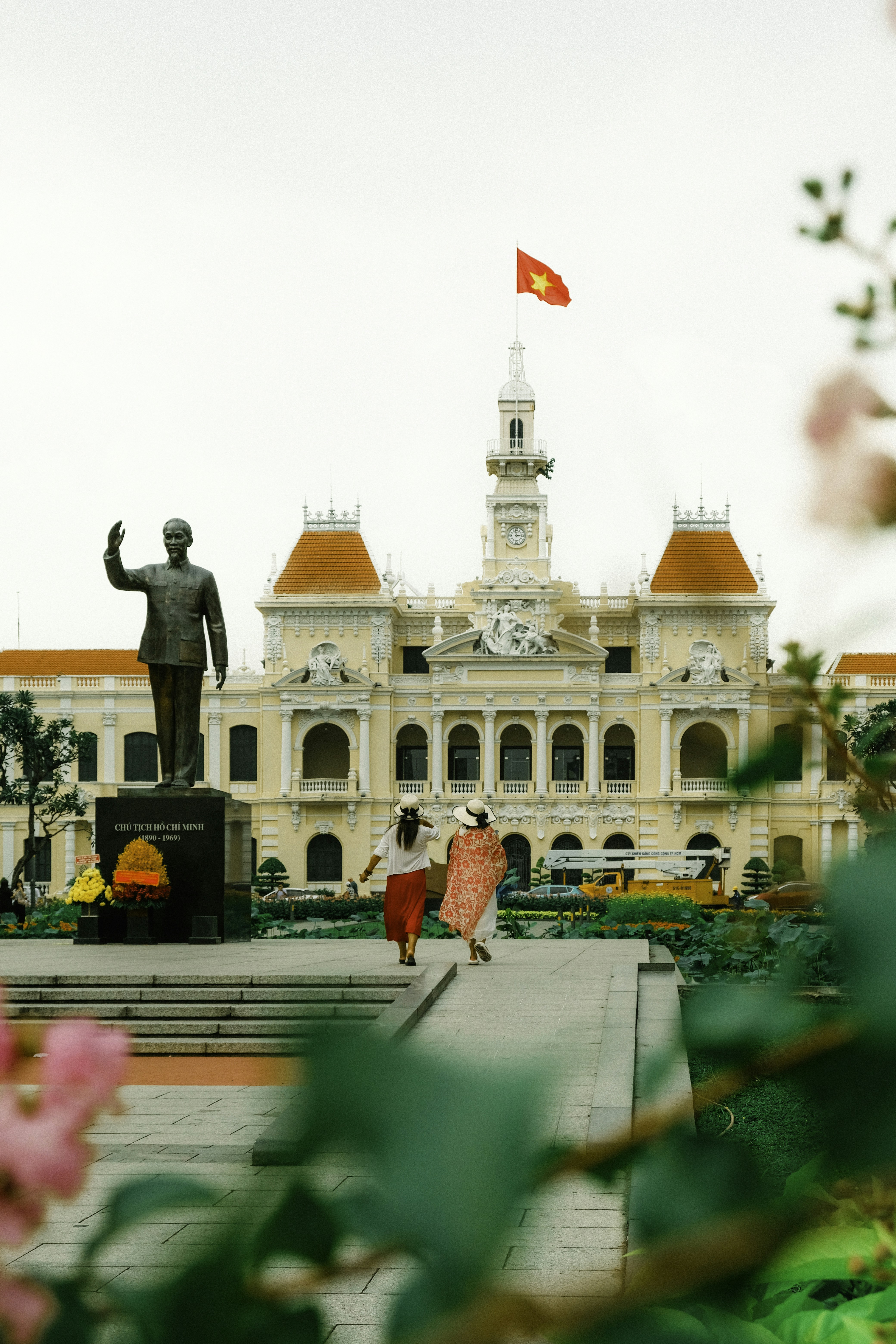 Two women walk along a paved plaza before a grand colonial building with a central clock tower and a red flag. A bronze statue stands to the left, while foreground greenery softly frames the scene.
