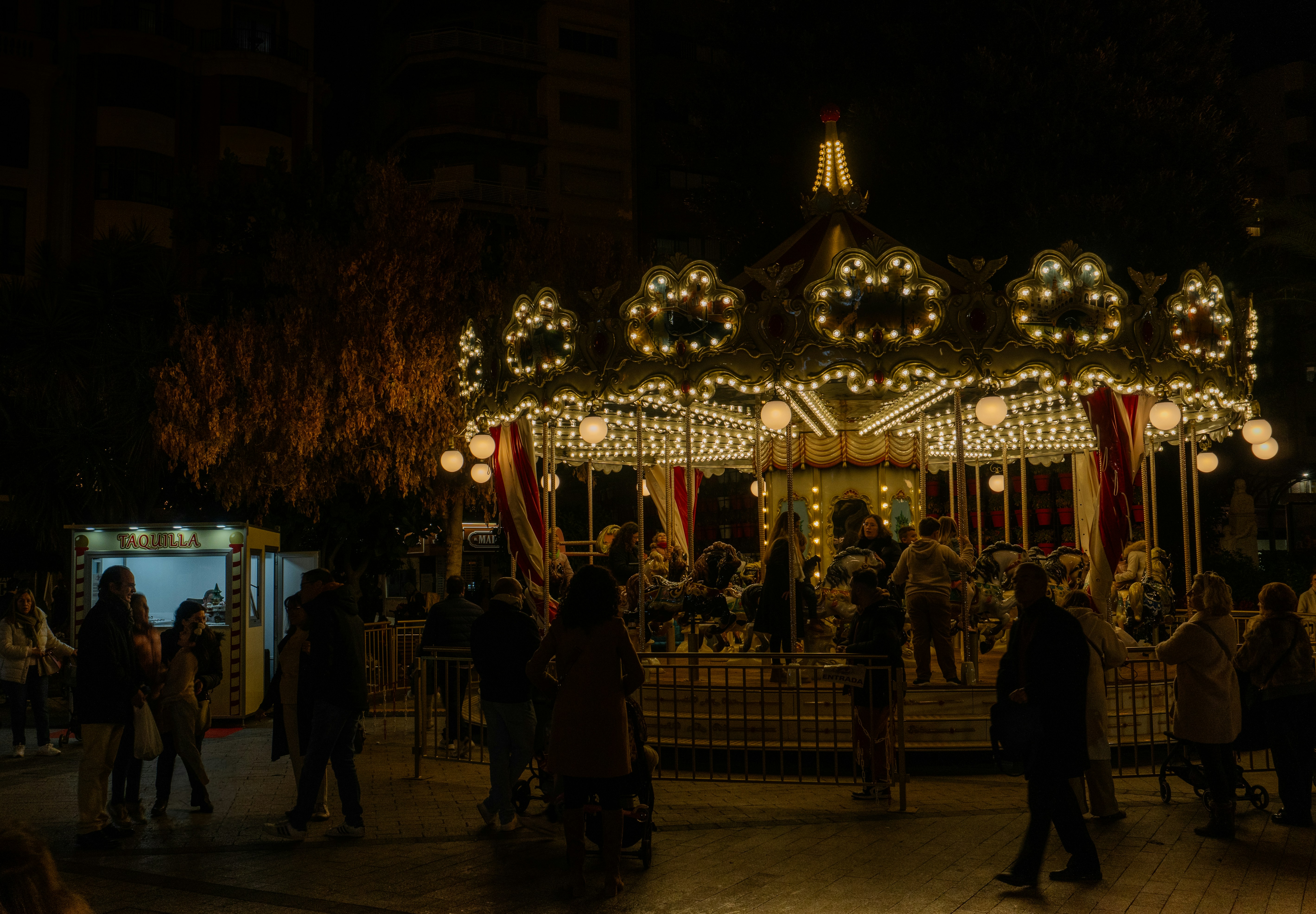 Illuminated carousel in a dimly lit urban square, surrounded by people enjoying the night.