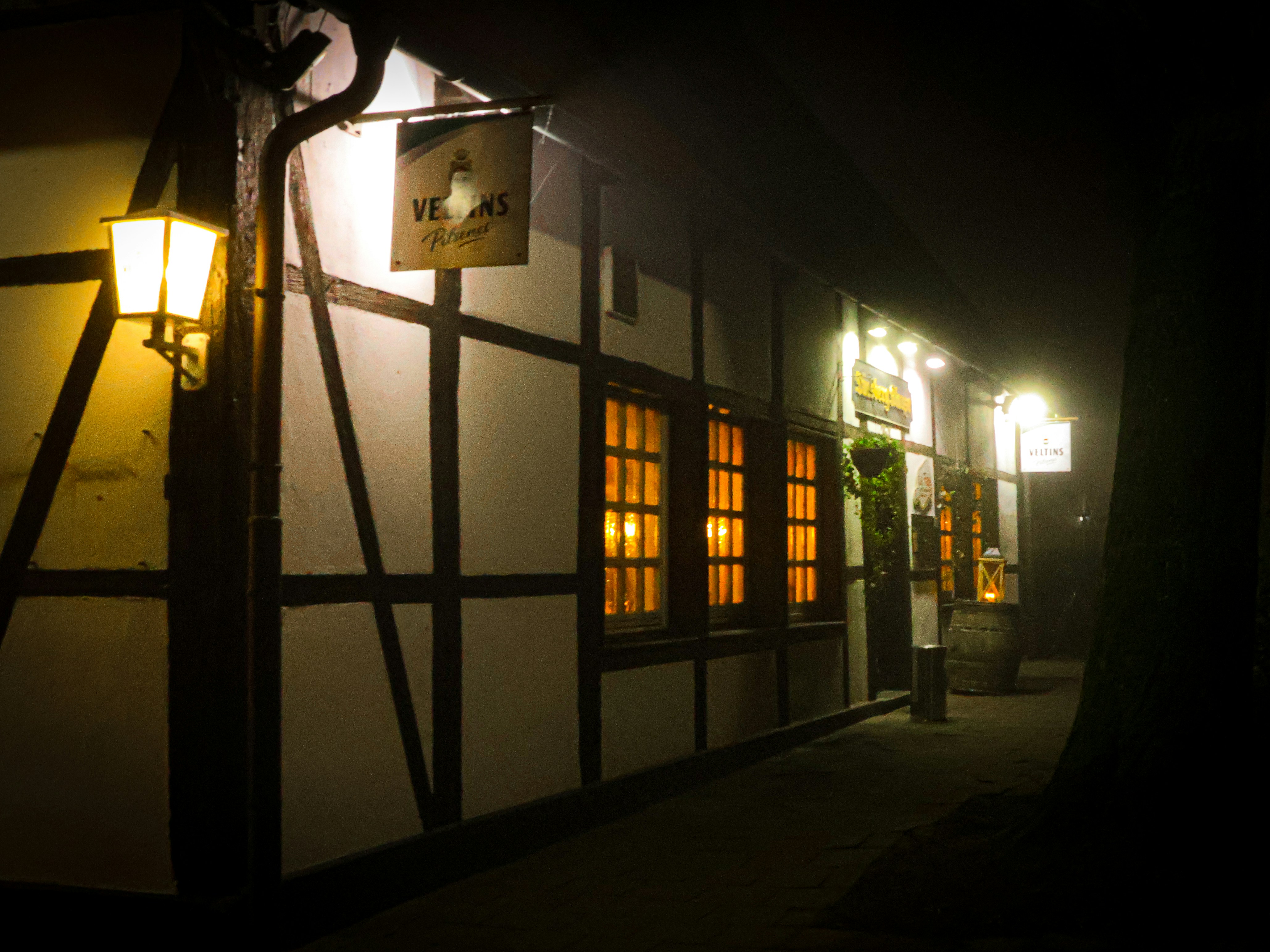 Rustic tavern with warm lantern glow on a foggy night, highlighting textured wood and white walls. Mist enhances the cozy, nostalgic atmosphere.