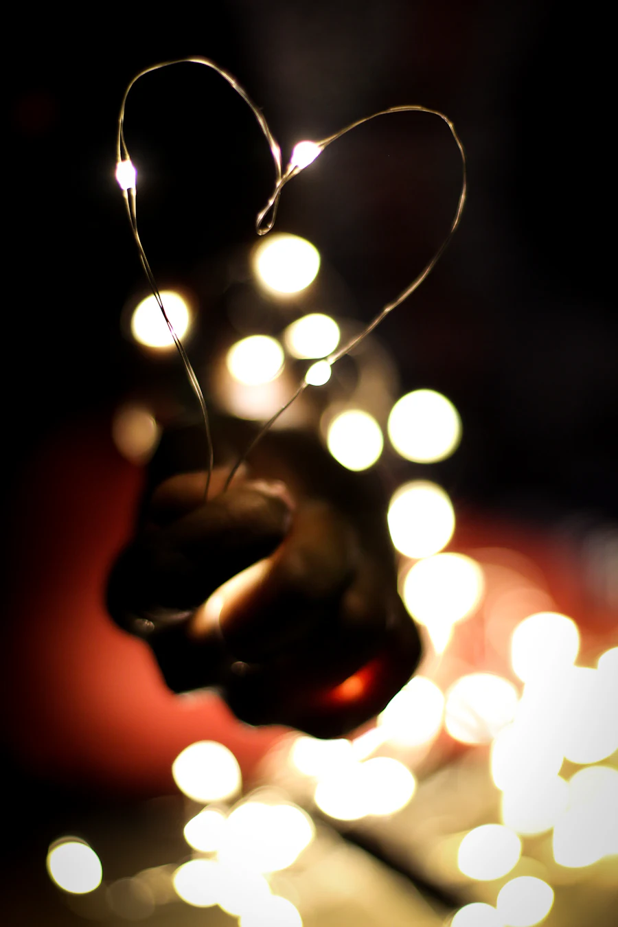 A hand holds a wire heart ornament surrounded by soft warm fairy lights against a dark background. The scene has a dreamy, out-of-focus glow with red tones near the bottom.