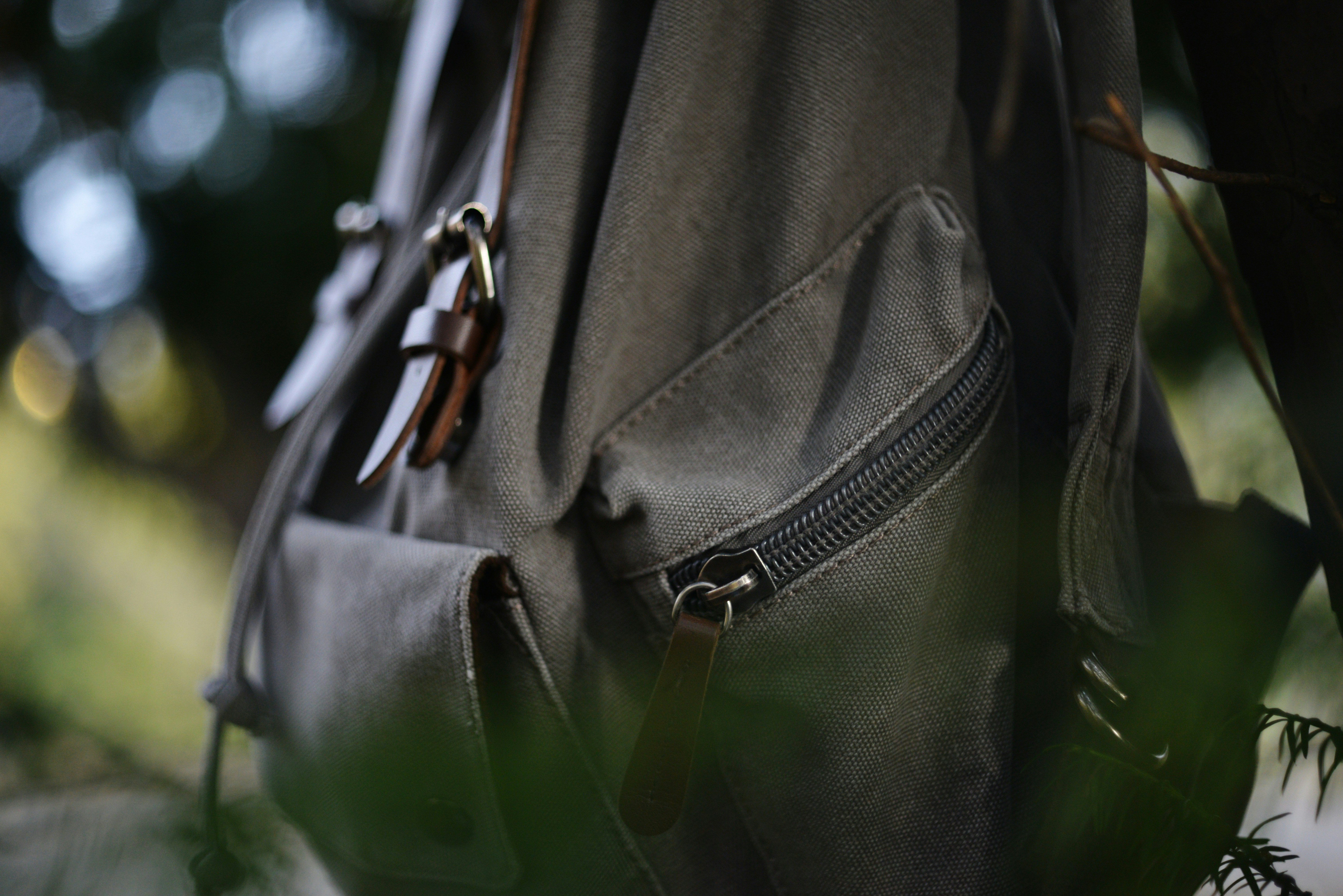Close-up of a gray backpack with leather accents, showcasing its pockets and zippers against a blurred natural background.