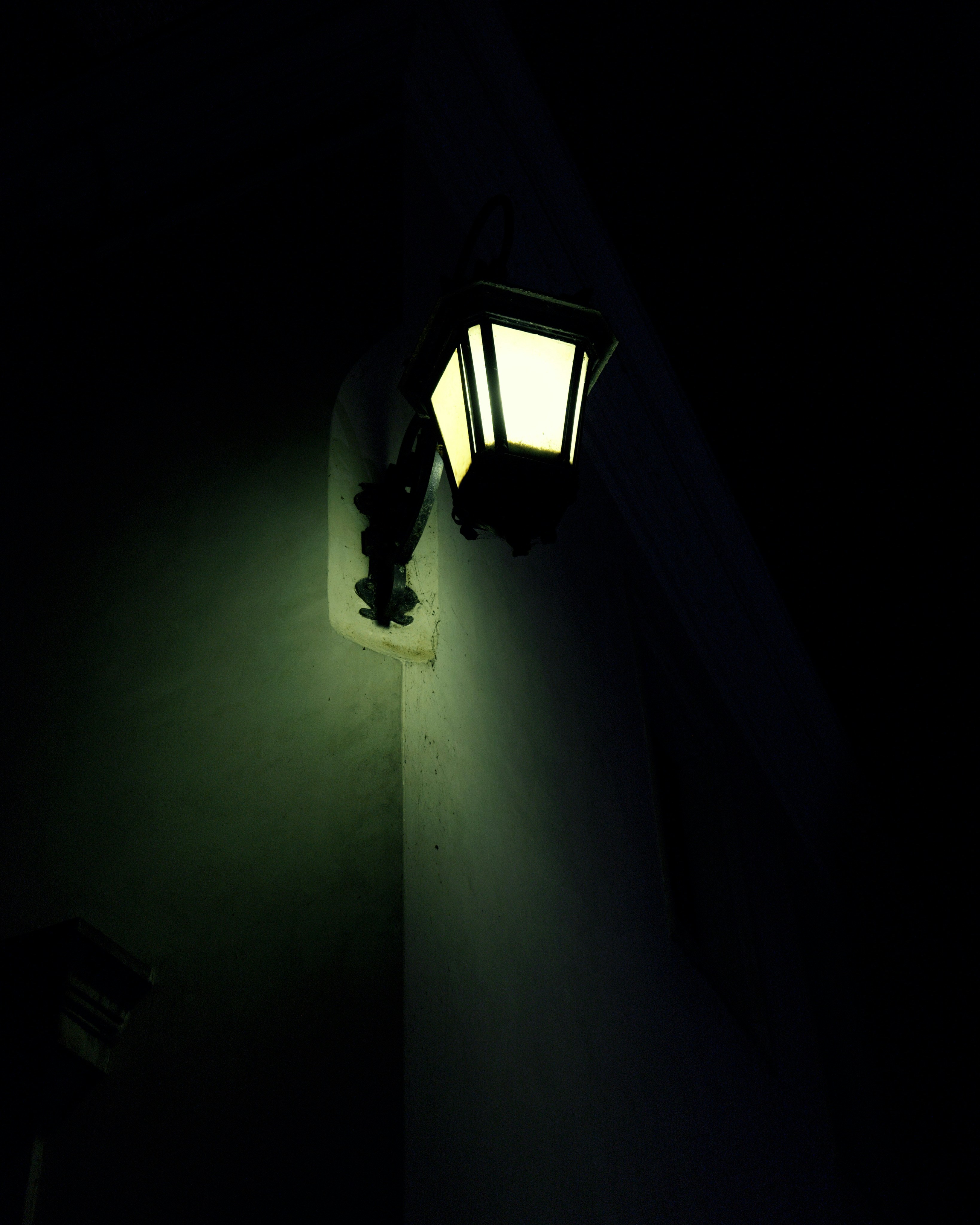 A single wall-mounted lantern casts a muted green glow on a textured facade, leaving most of the scene in deep shadow.