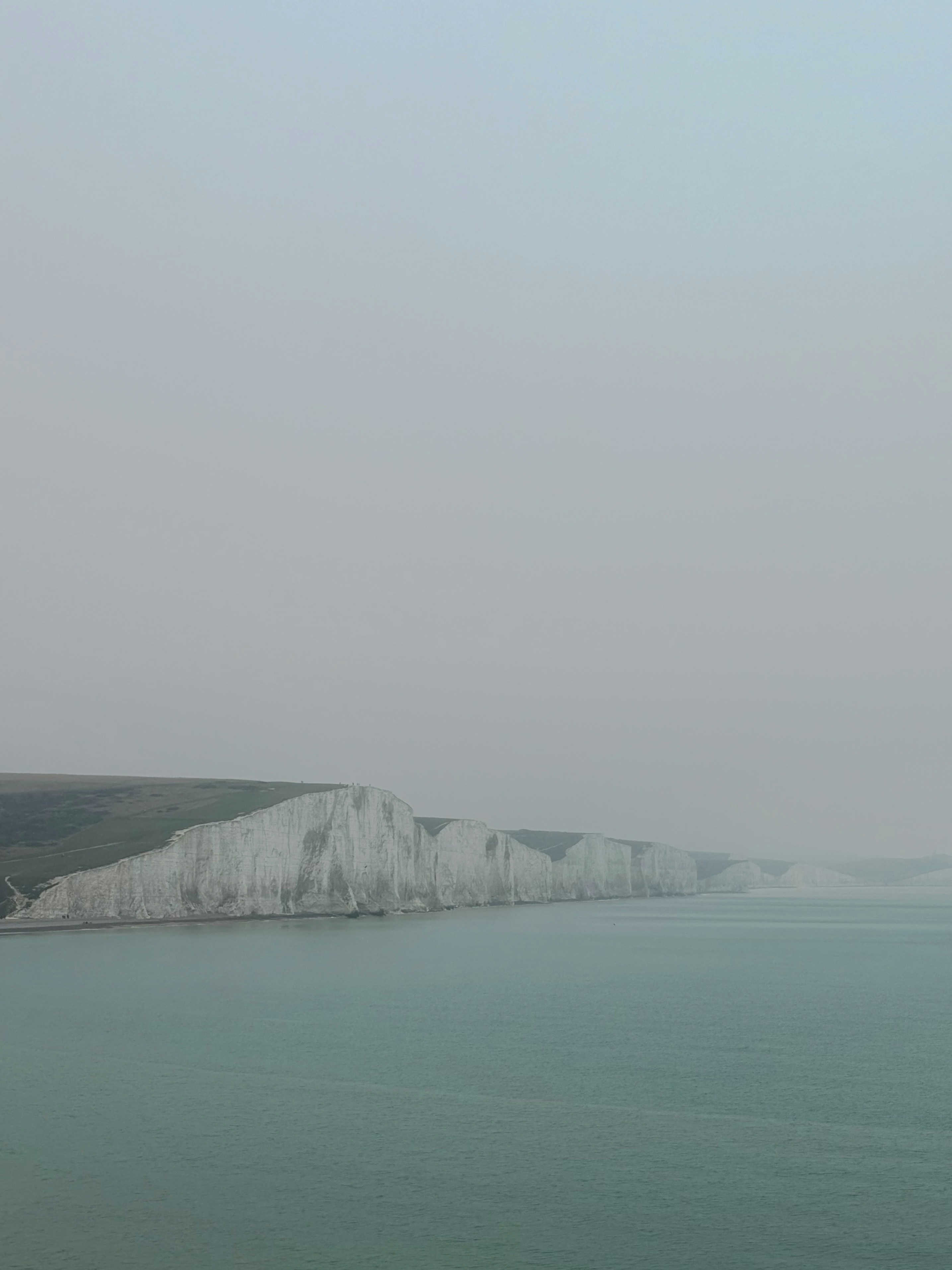 Chalk cliffs stretch along a calm turquoise sea under a hazy sky.