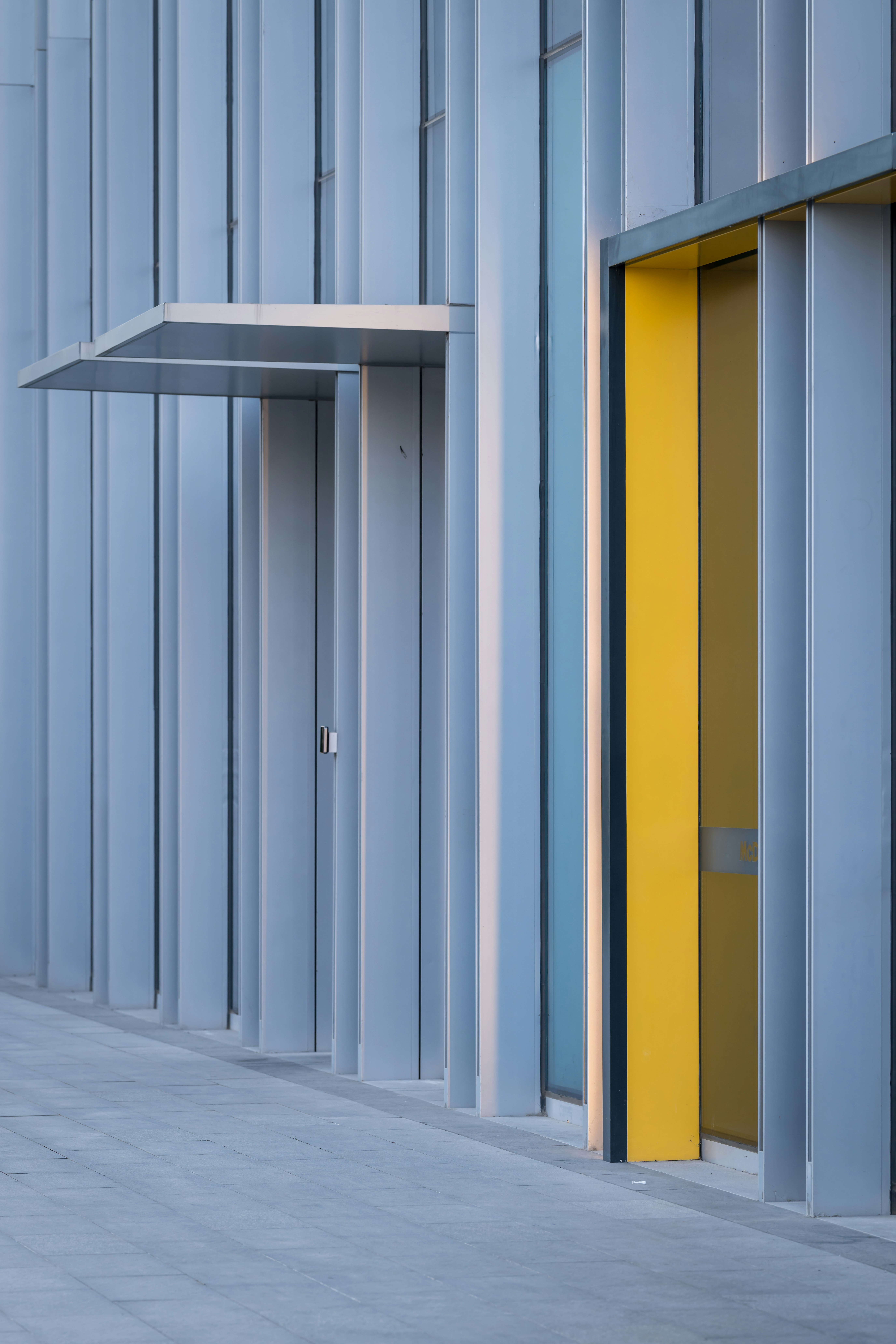 Blue vertical metal cladding frames a bold yellow doorway, creating strong color contrast. The composition emphasizes modern architectural geometry and rhythm.
