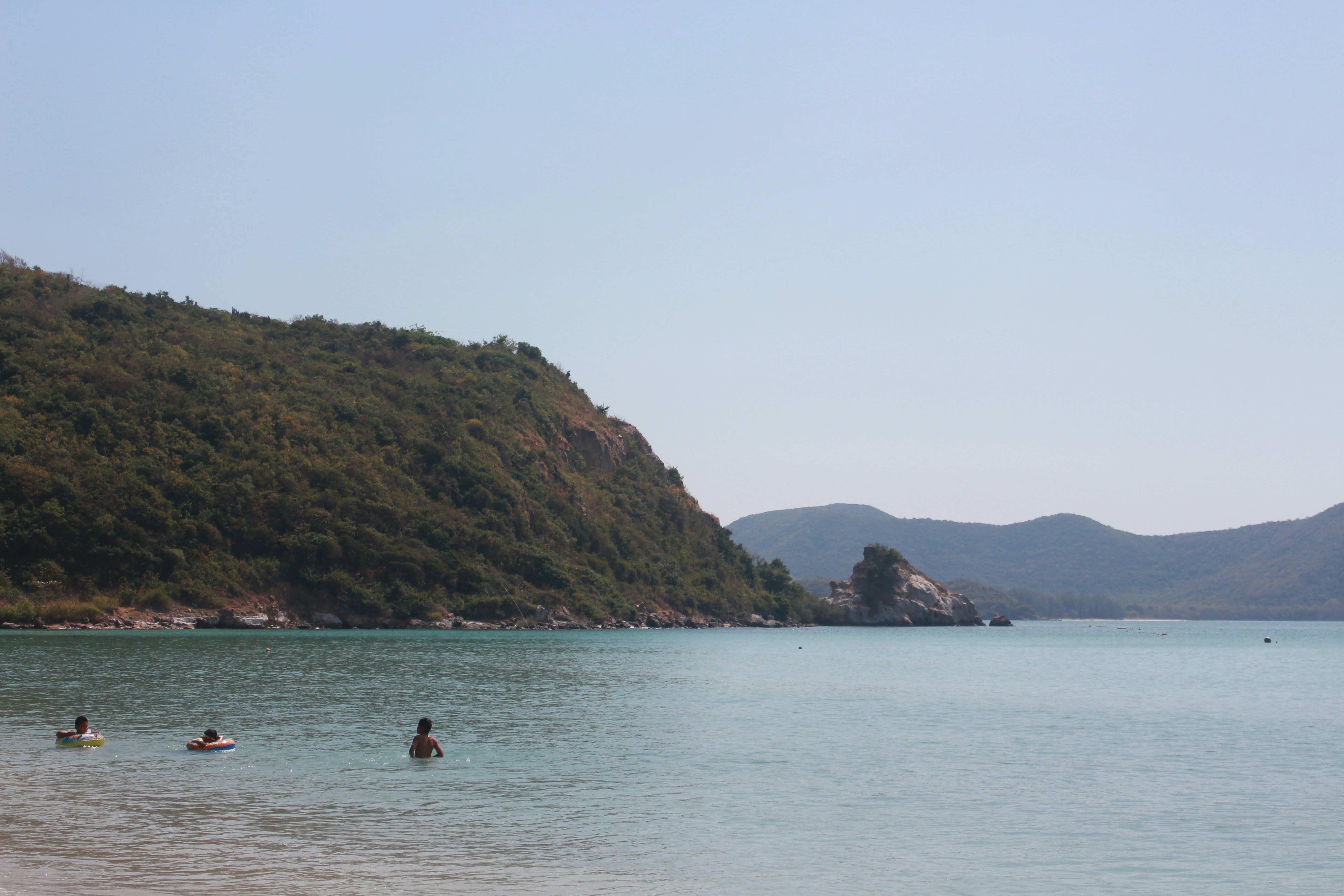 Swimmers enjoy the calm turquoise waters near lush green hills under a clear sky.