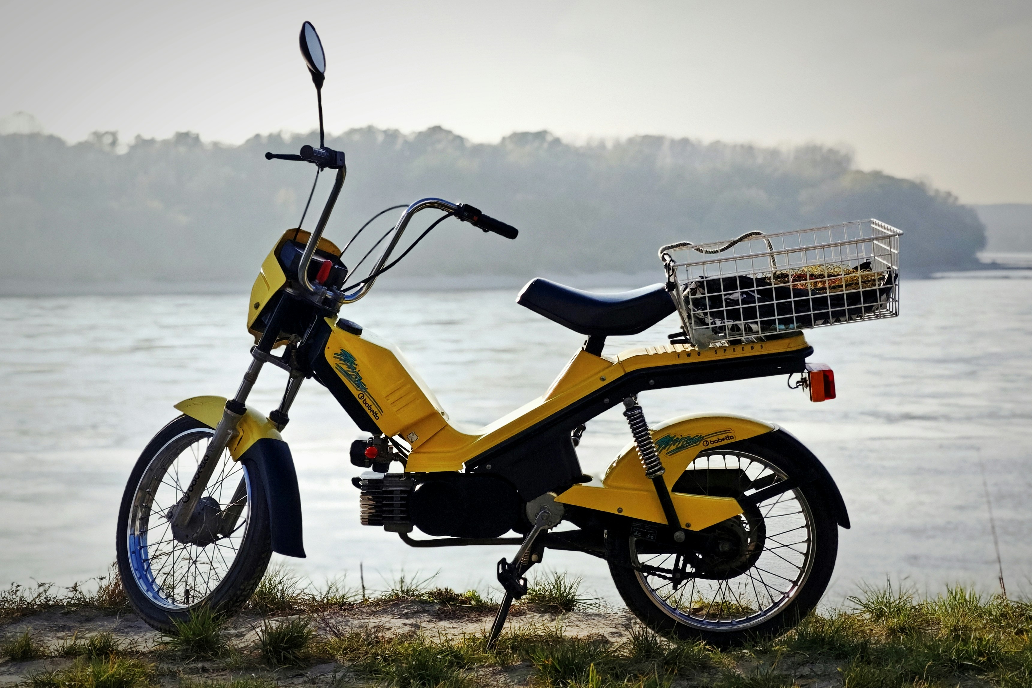 A yellow moped with a rear basket rests on grass beside a calm river, with a hazy tree line in the background.