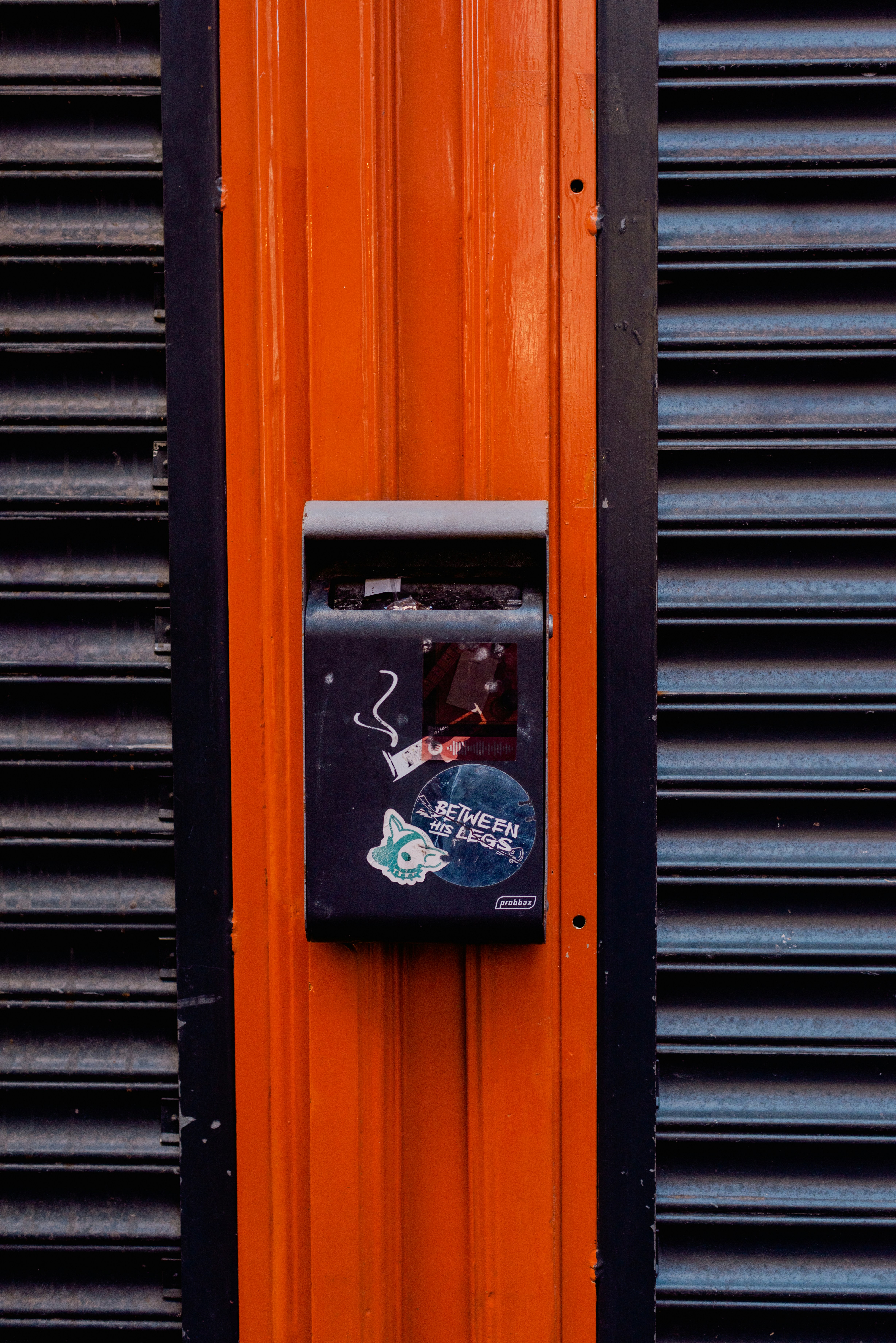 Between the Grates: A Stickered Mailbox Framed by Orange Steel