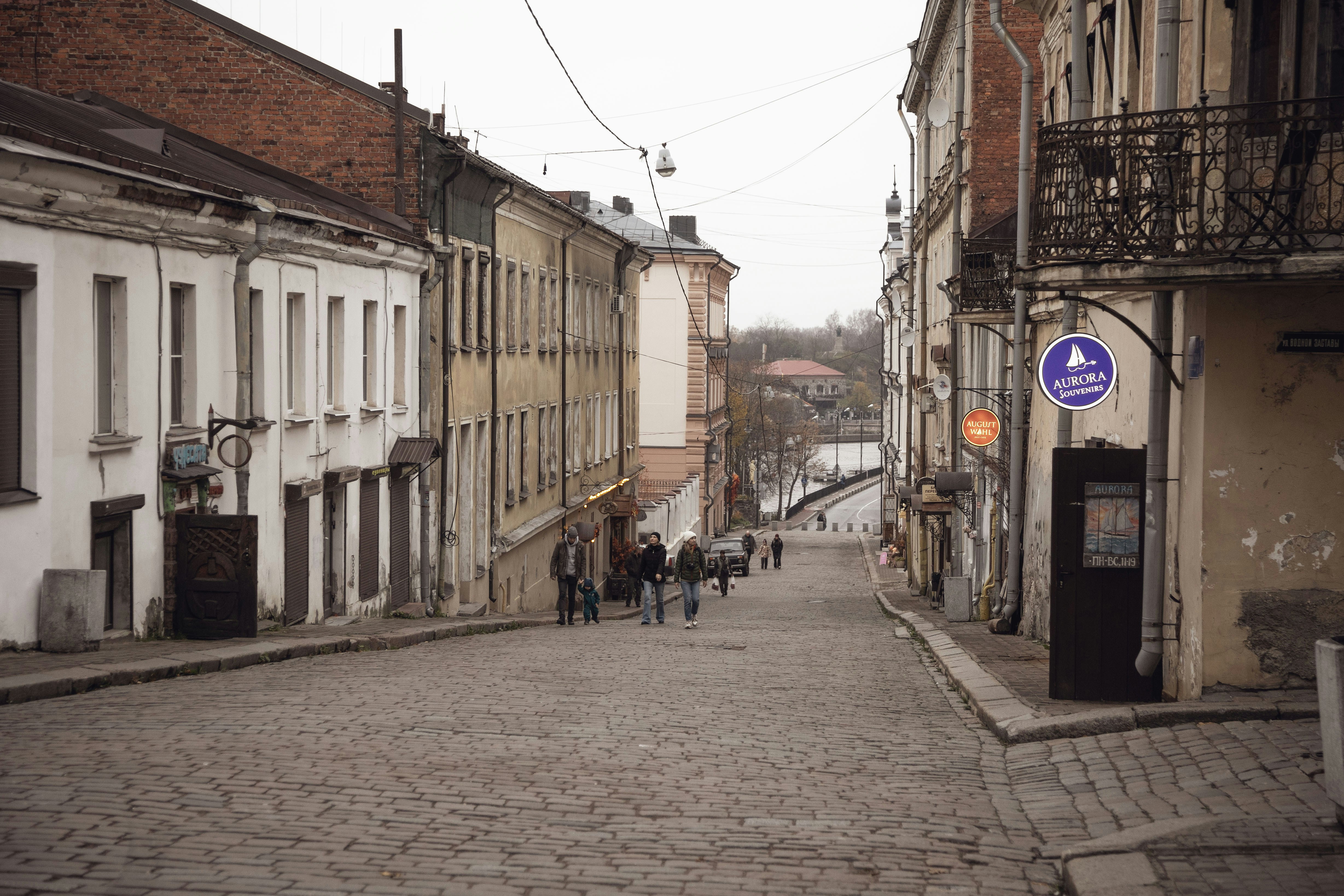 Cobblestone street flanked by aged buildings leading to a distant river under an overcast sky.