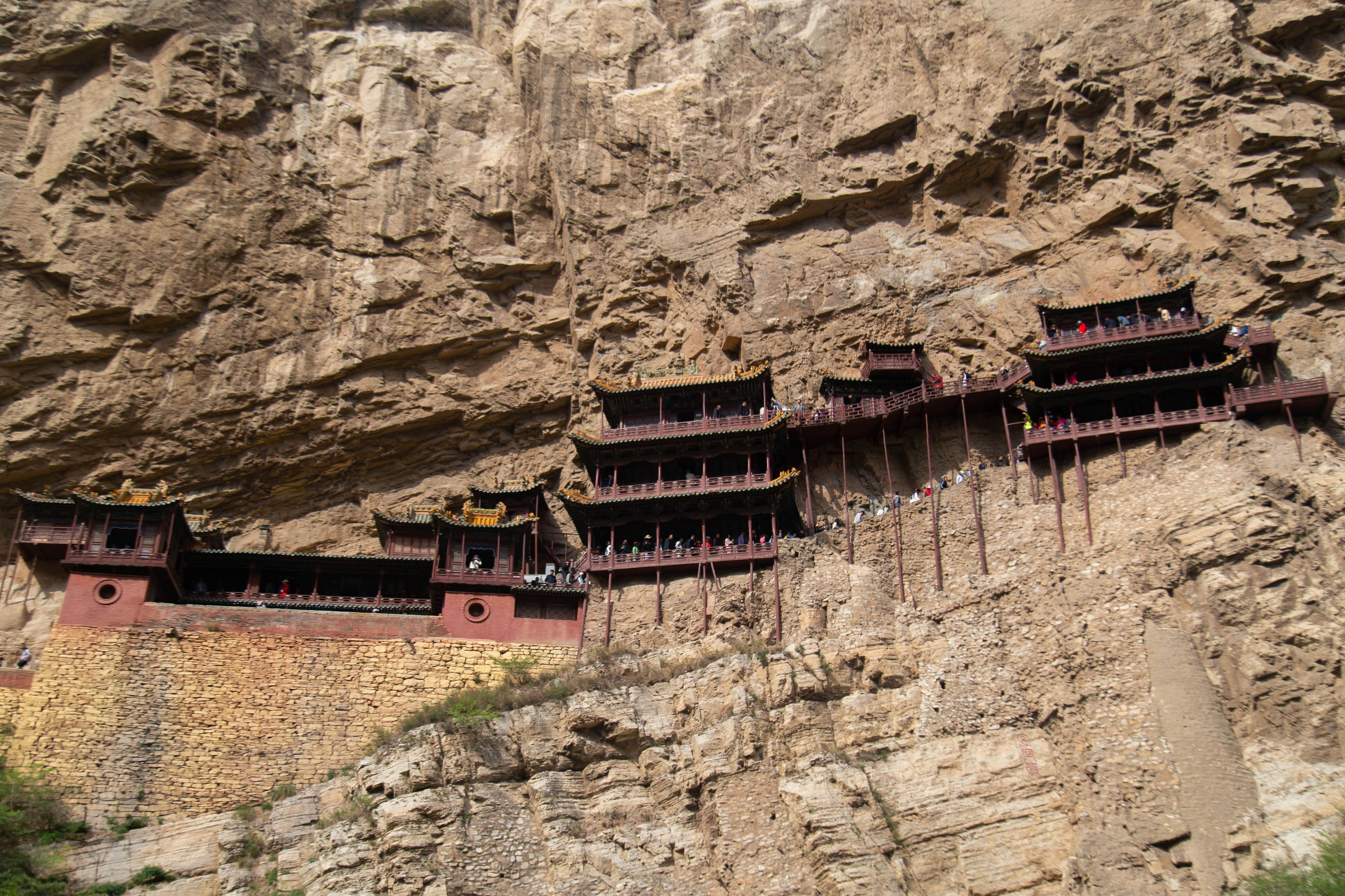 Ancient wooden temples cling to a sheer cliff face under a clear sky.