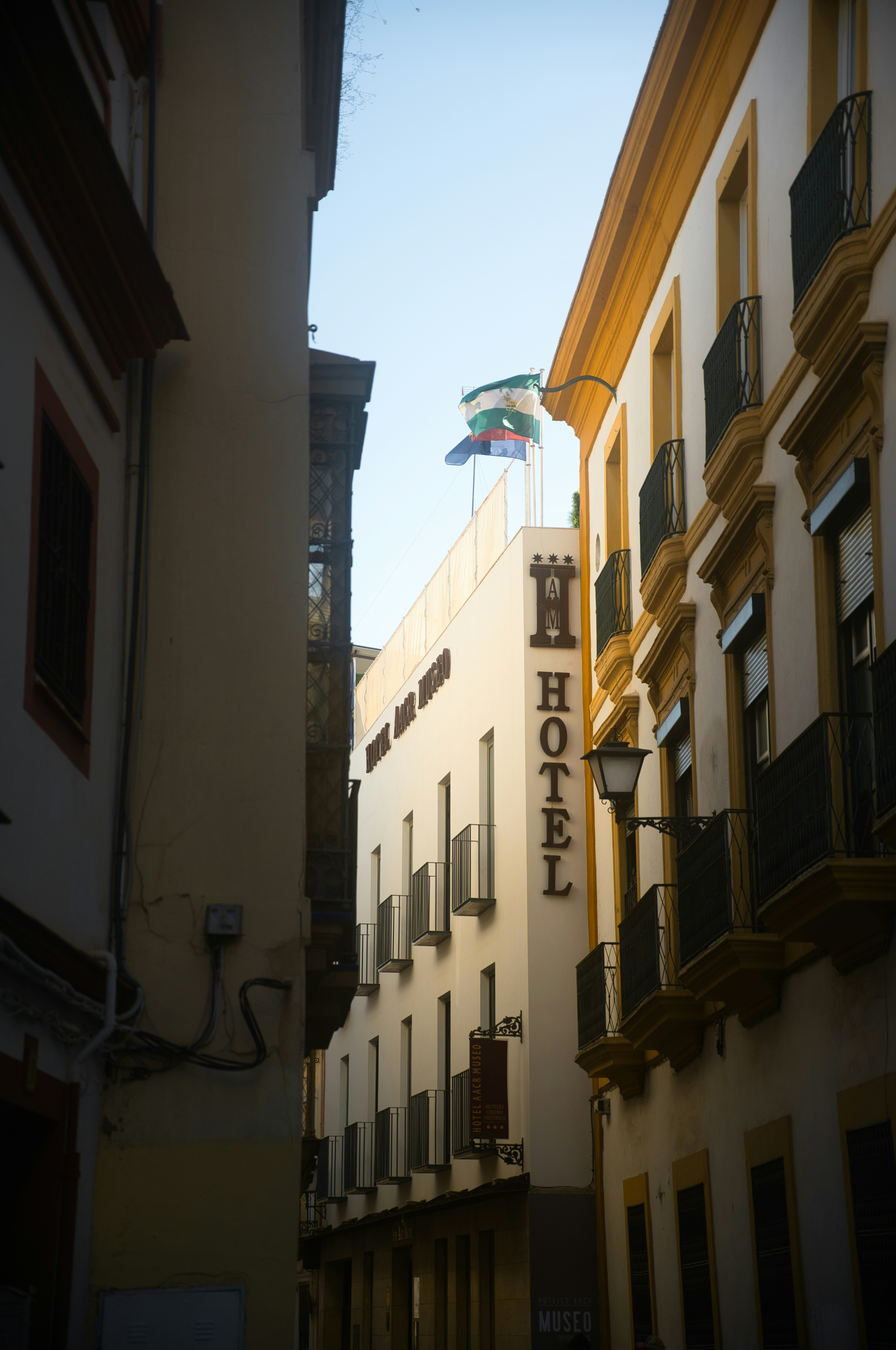 Sunlit Calle: A Narrow Alley Framed by a Vertical Hotel Sign and Fluttering Flags