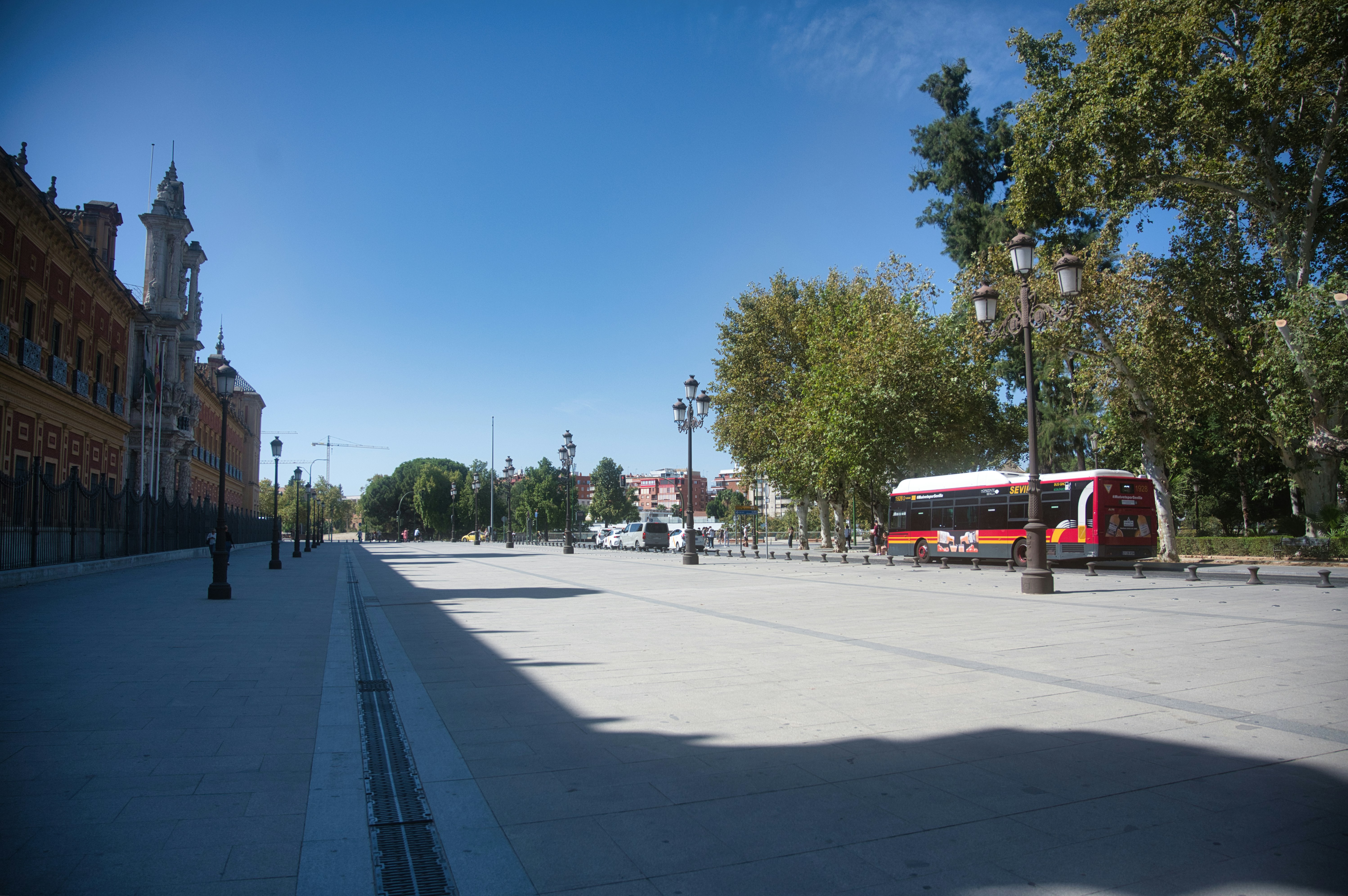 Wide street with a red bus parked under clear blue skies, flanked by historic buildings and lush trees.