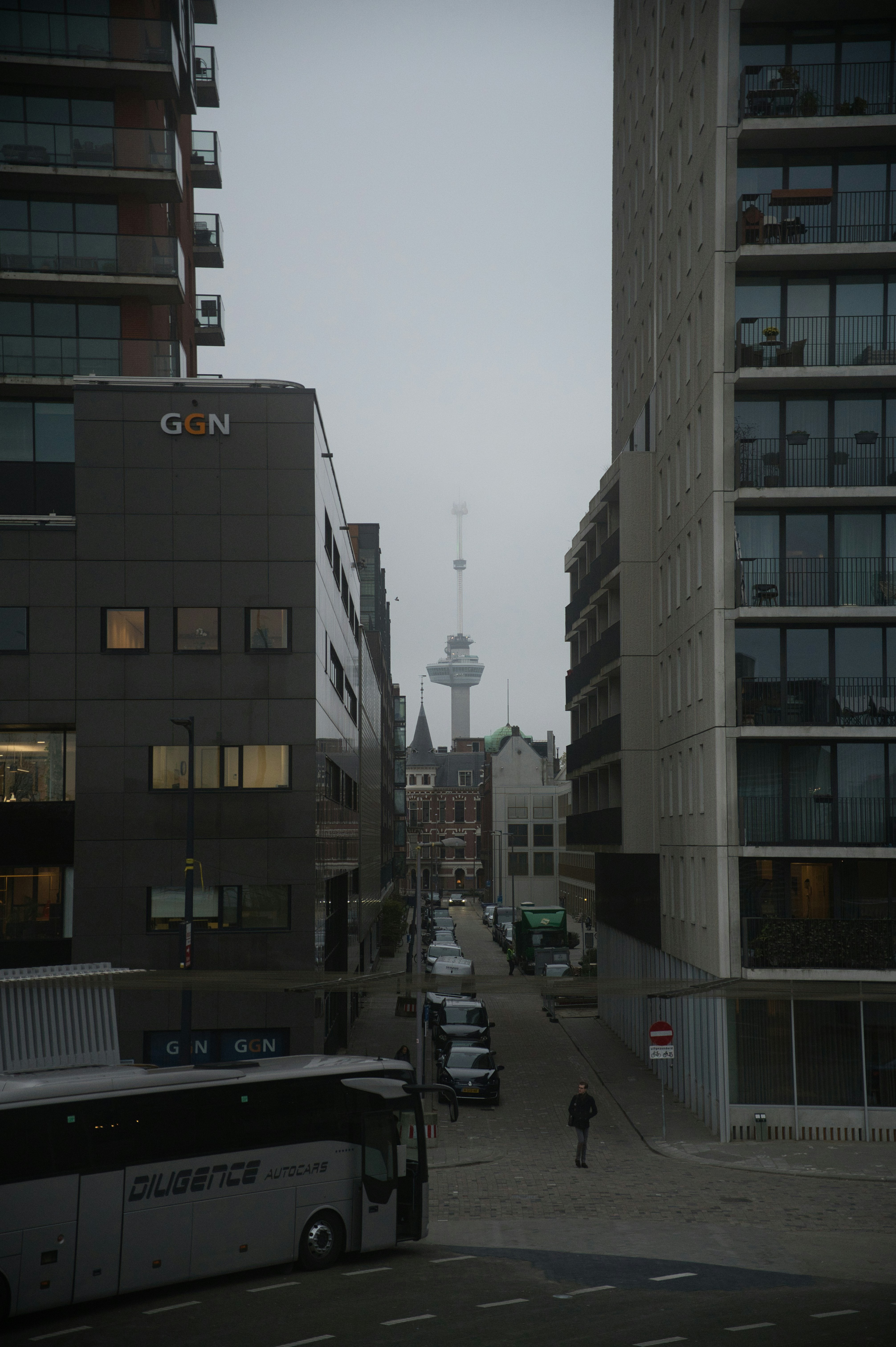 A city street framed by modern buildings, revealing a distant tower shrouded in fog. The scene captures a blend of urban architecture and atmospheric mystery.