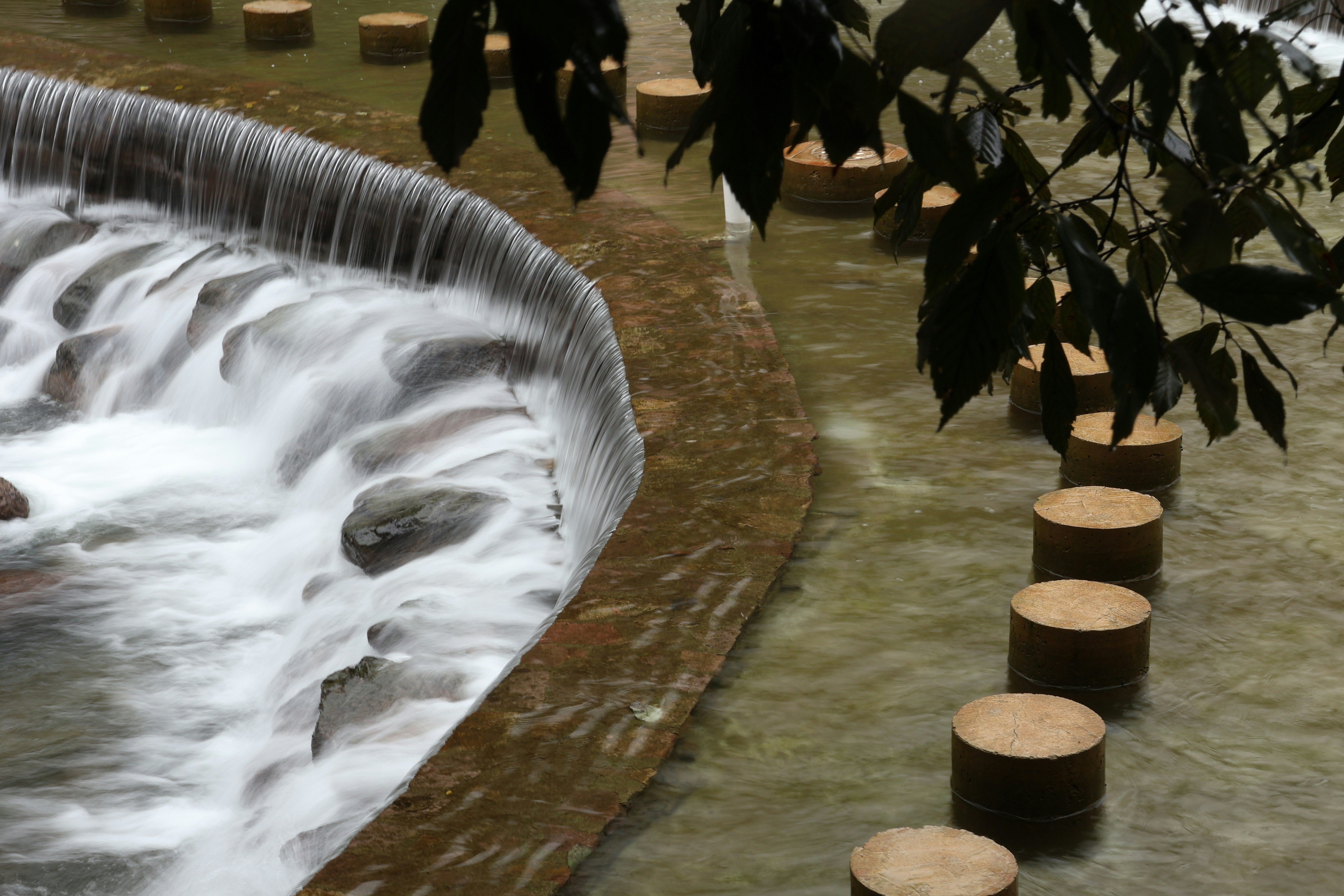 Curved stepping stones lead across a flowing stream beside a small waterfall, framed by overhanging leaves.