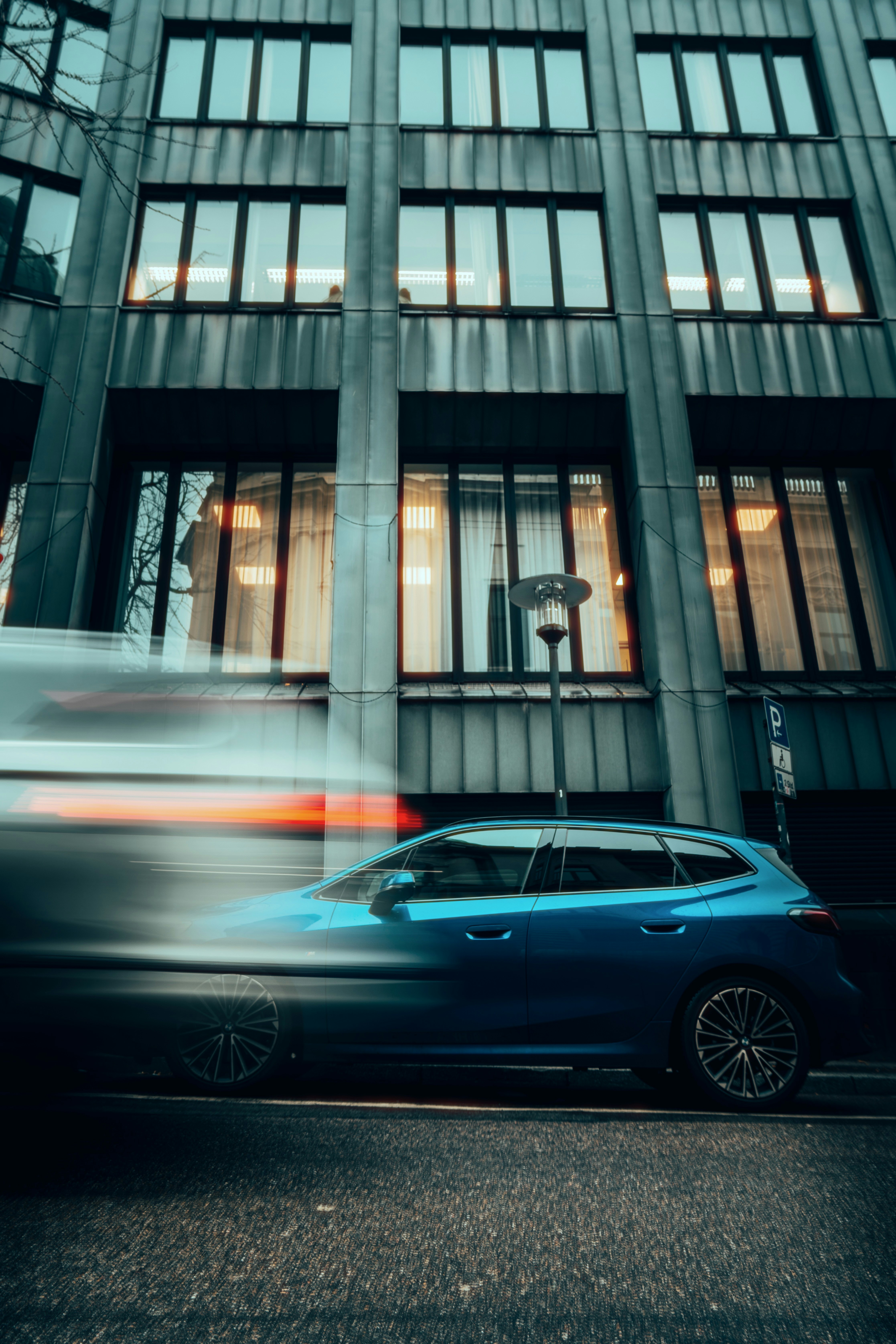 Blurred car speeding past a stationary blue vehicle with a modern building facade in the background.