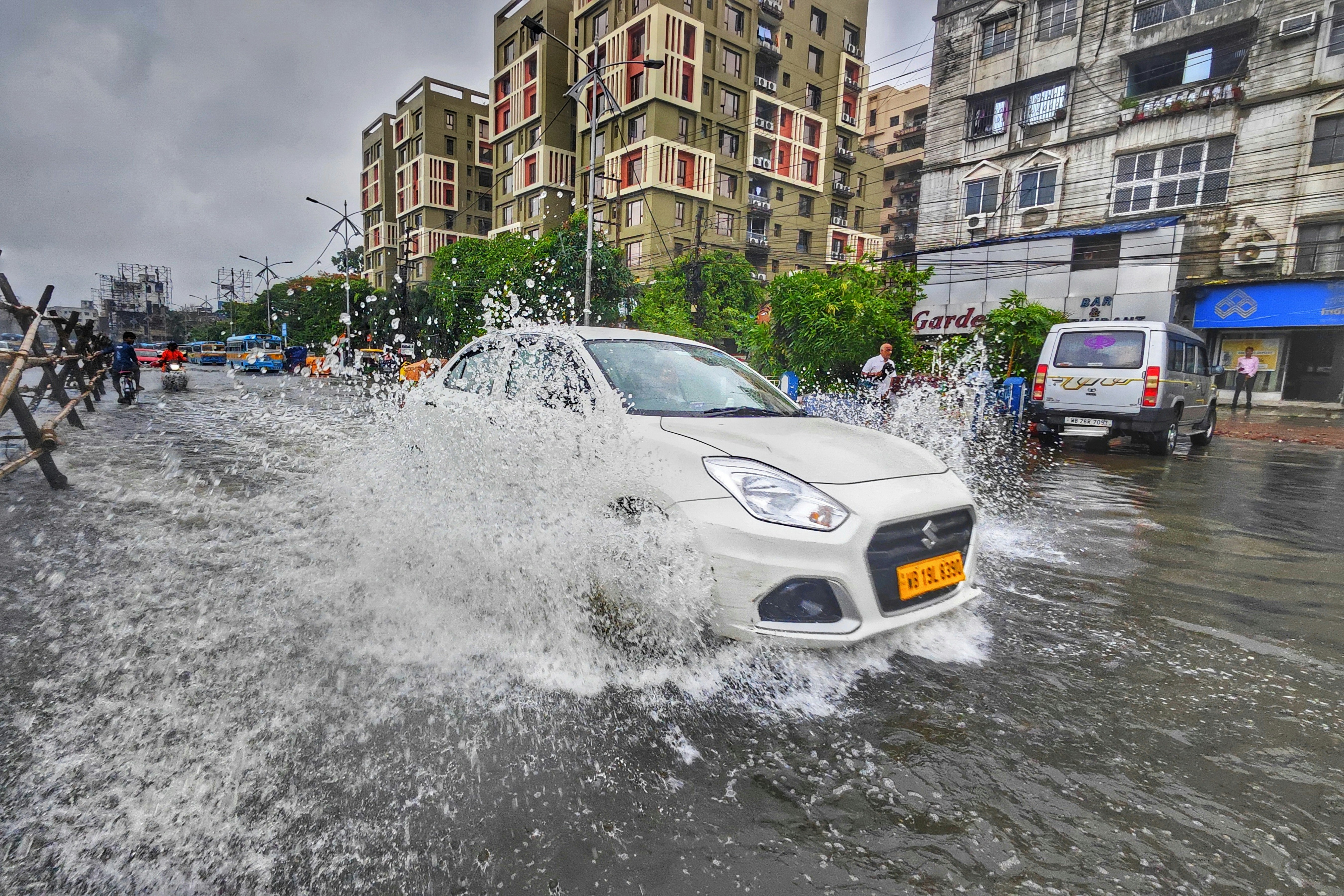Photograph of a white sedan plowing through floodwater on a busy city street, spraying water across the foreground. Buildings and pedestrians frame the scene.