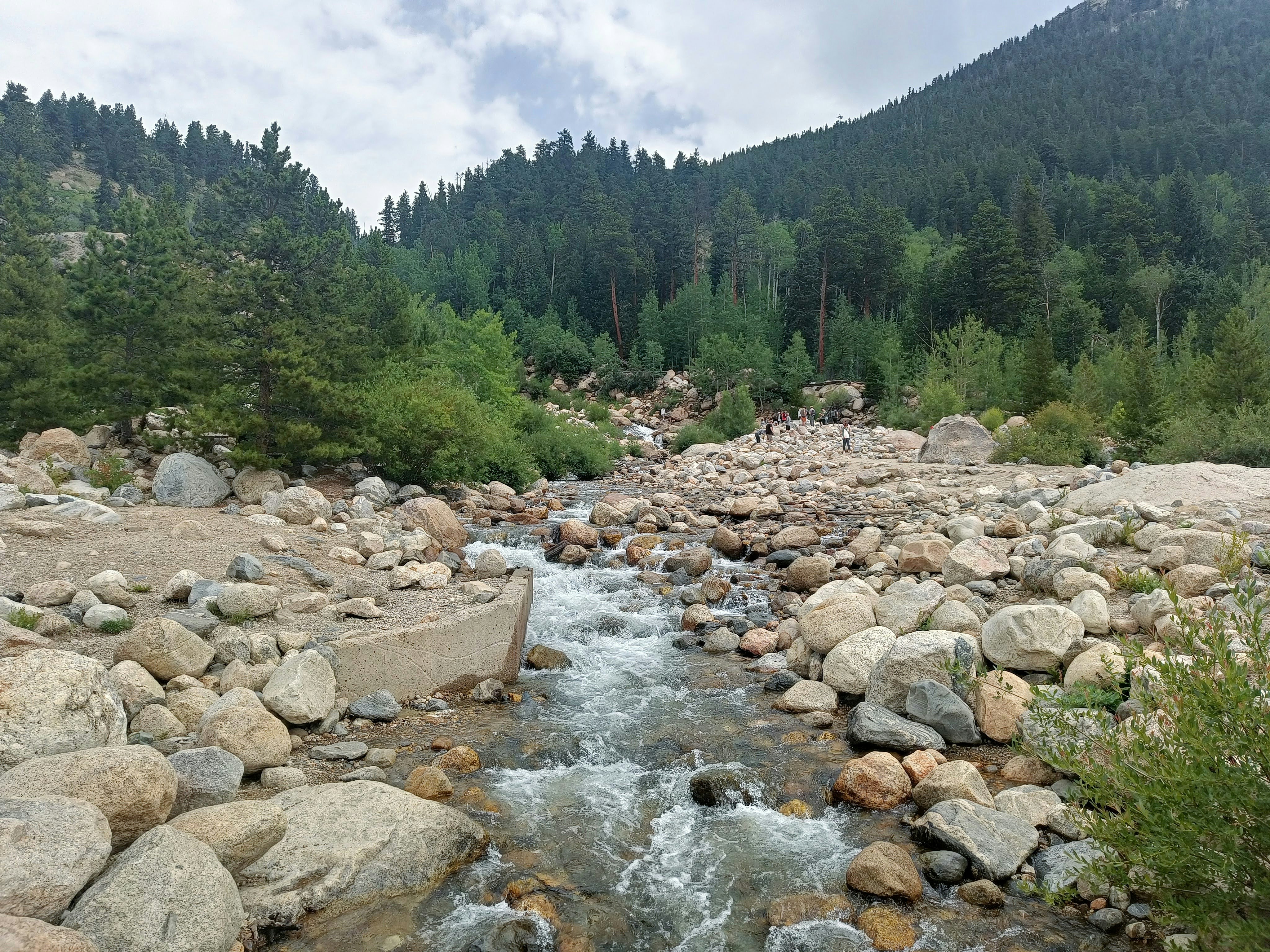 A rocky riverbed flows through a lush green landscape, surrounded by towering mountains and scattered boulders. People can be seen exploring the serene environment.