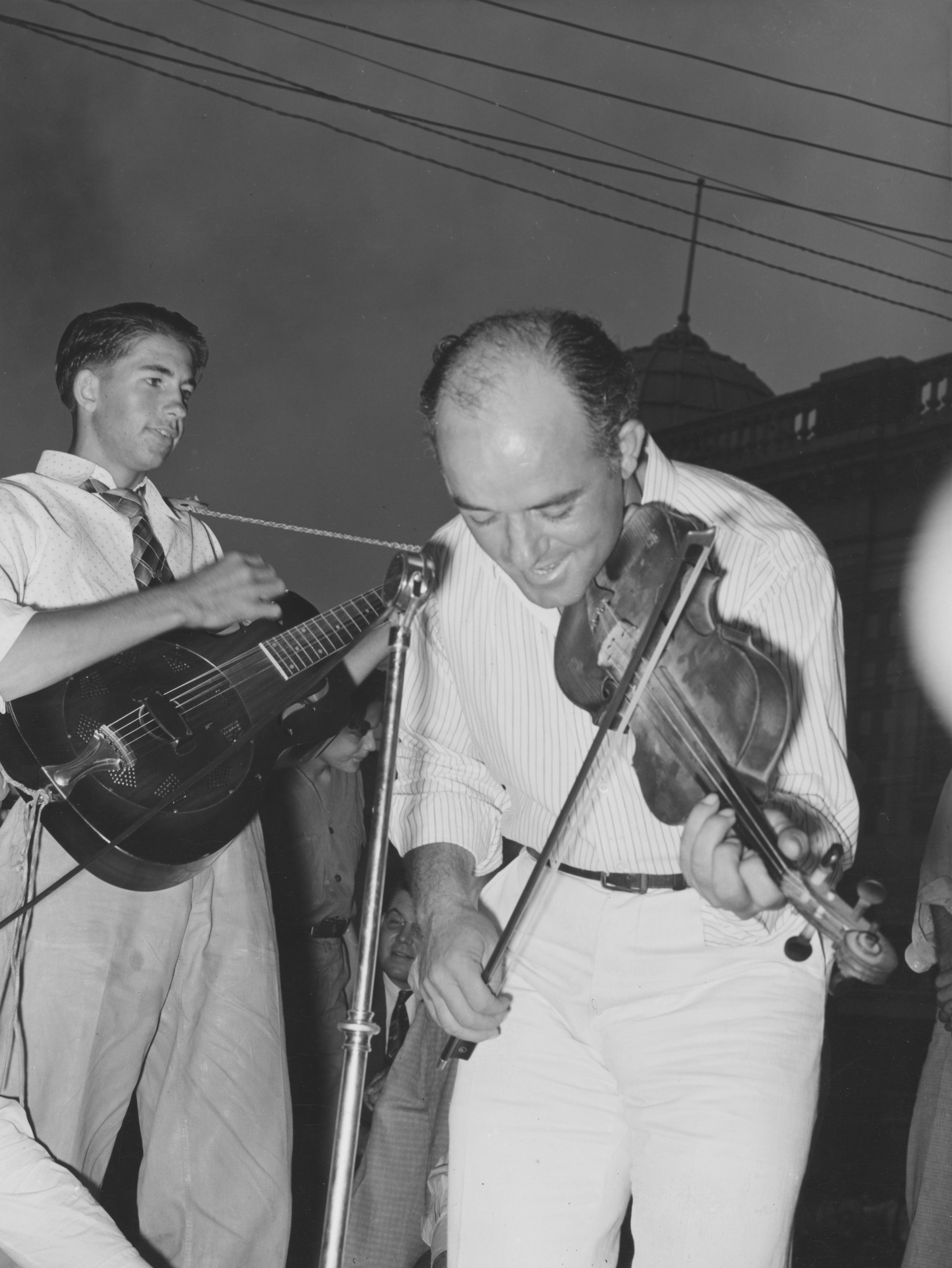 A candid street performance featuring a violinist leaning into his instrument while a guitarist accompanies nearby under urban lights.