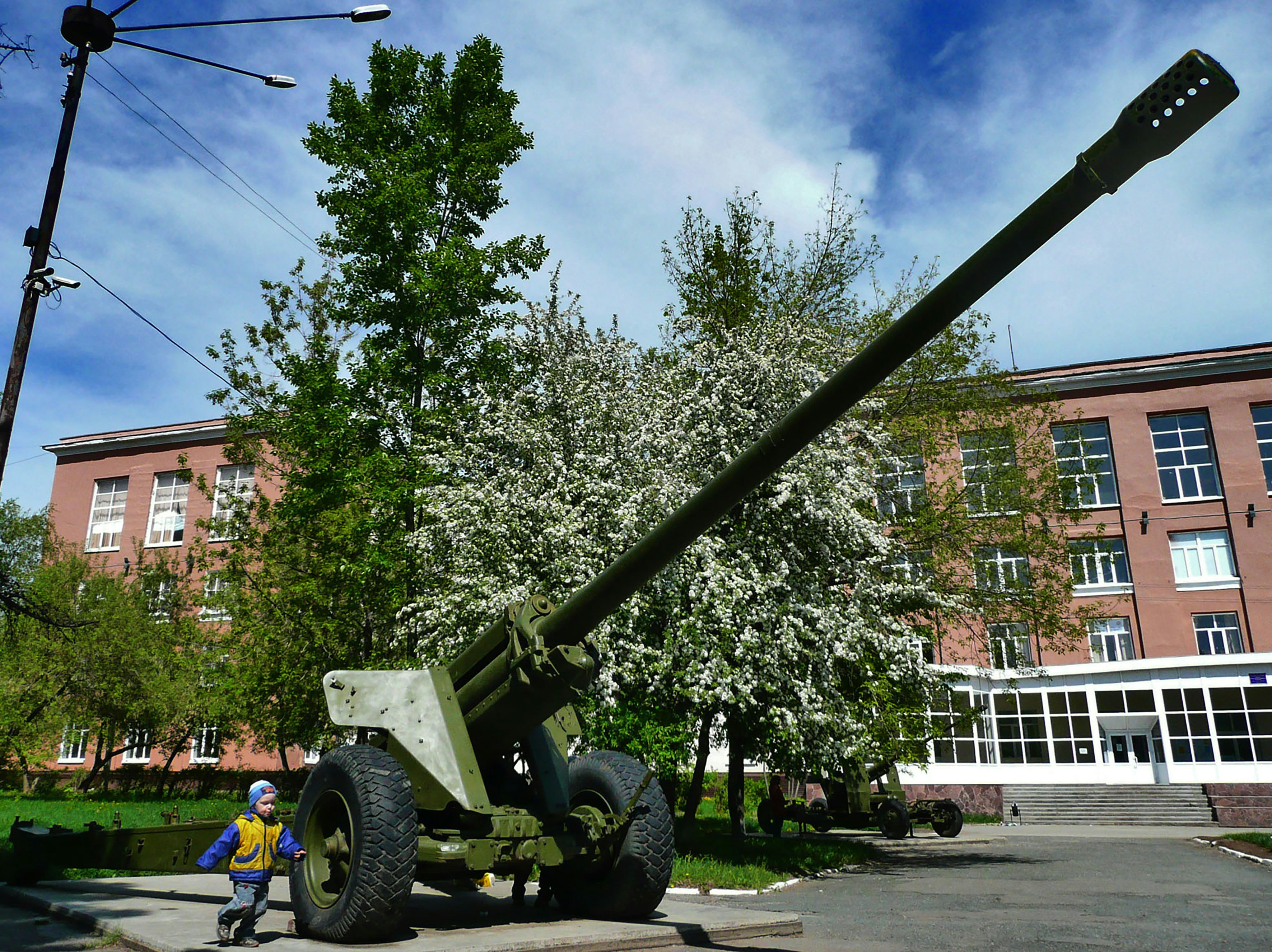 A historic field gun sits on a paved area beside a brick school building, framed by blooming trees under a bright sky.