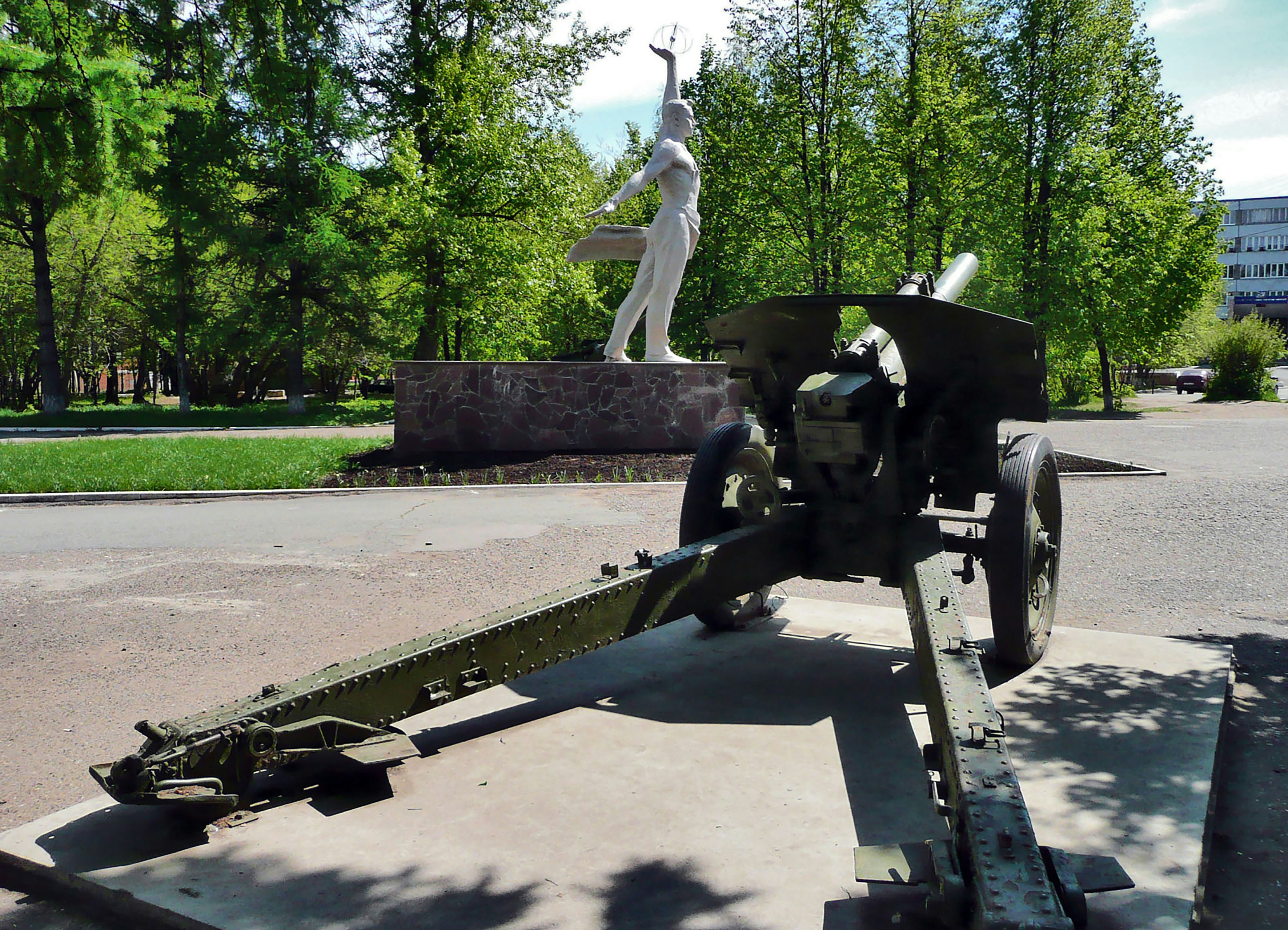 Artillery remains frame a plaza with a white statue in the background and green trees beyond; the cannon dominates the foreground, casting shadows on the pavement.