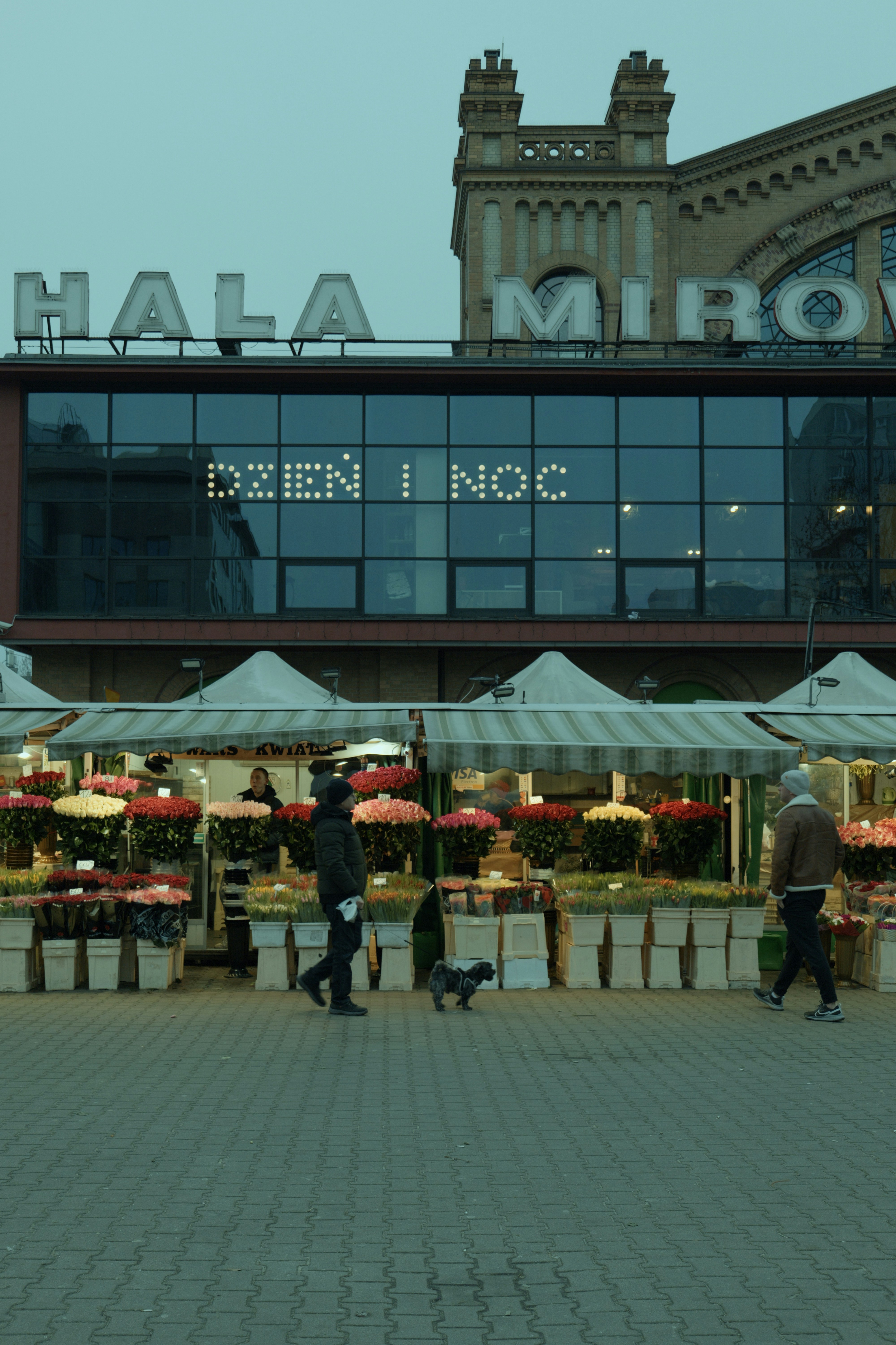 Dusk at Hala Mirowska: Glass Facade Framed by a Flower Market