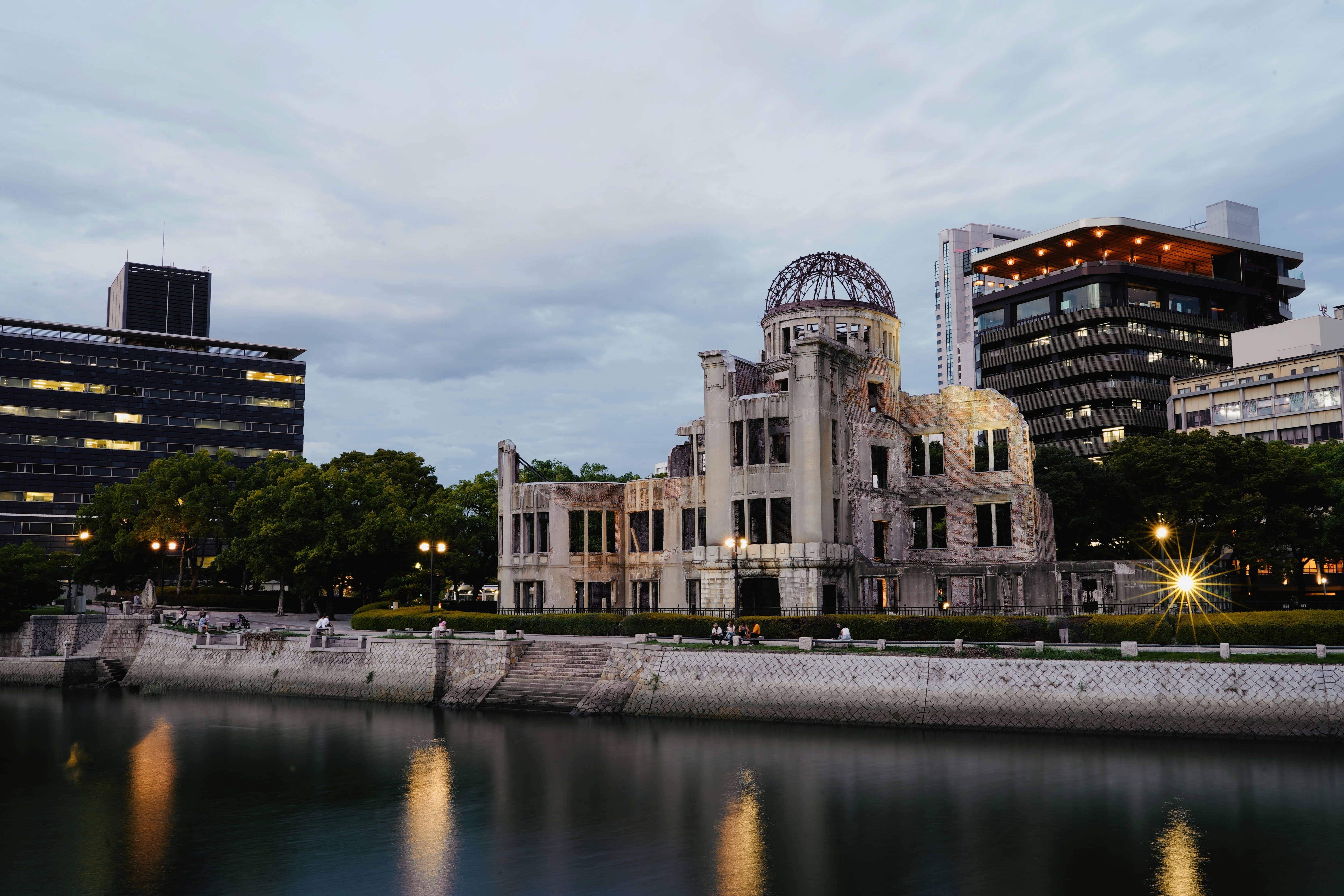 Hiroshima Peace Memorial with surrounding buildings by a calm river at dusk.