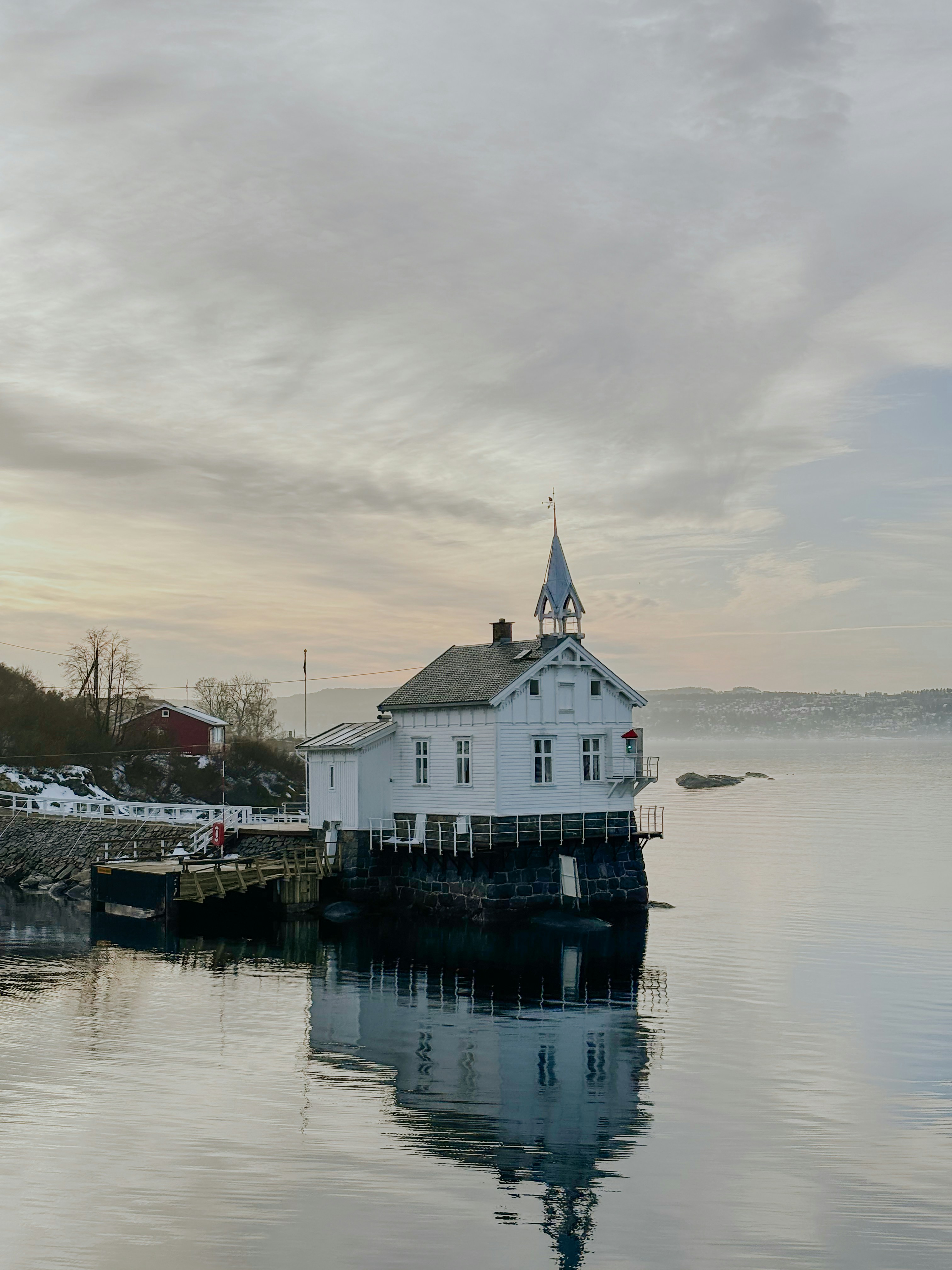 A white boathouse with a pointed steeple sits on a rocky foundation, connected by a wooden dock. The calm water mirrors the pastel sky above.
