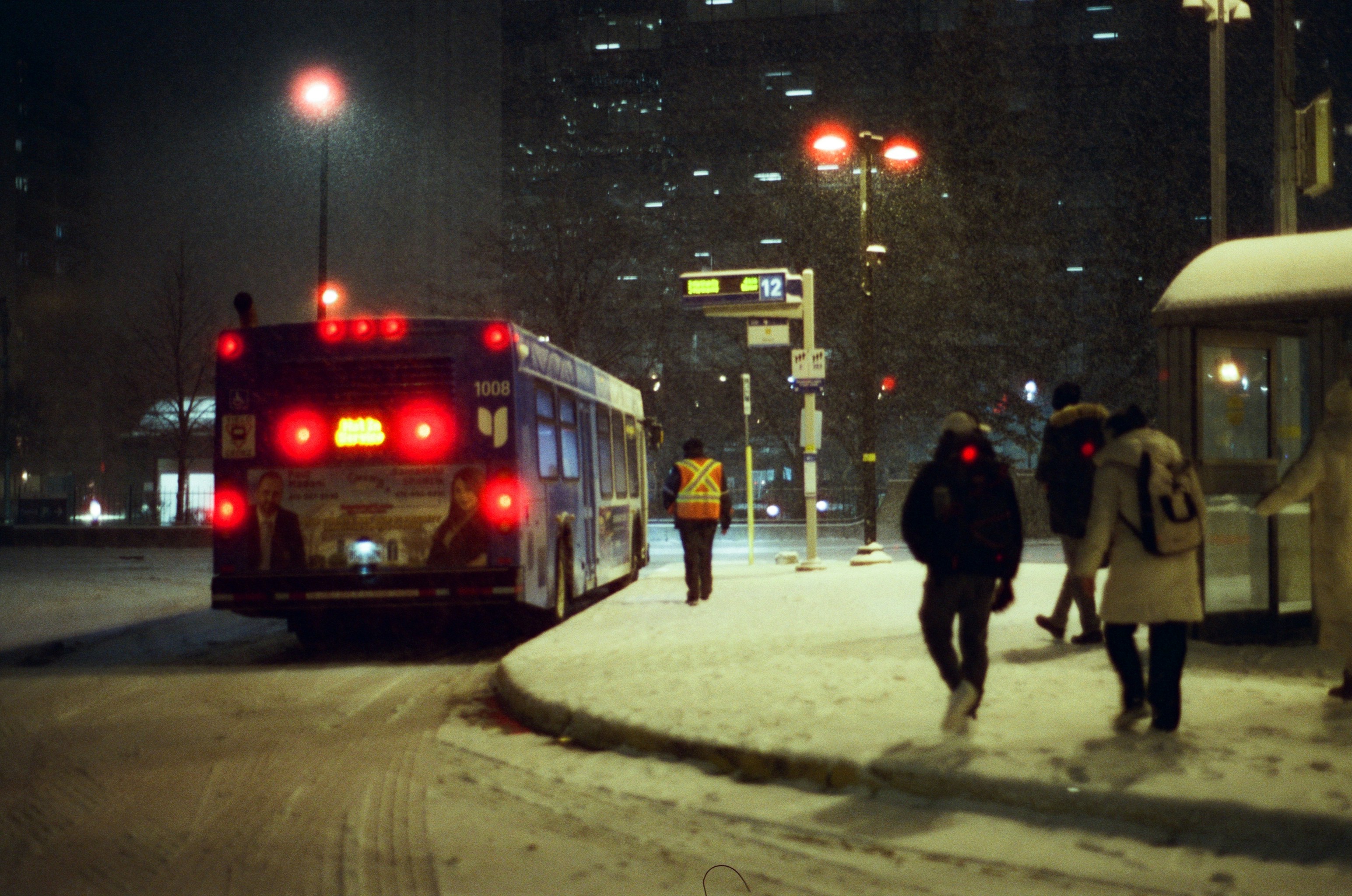 Bus halted at a snowy urban stop with passengers walking nearby under streetlights.