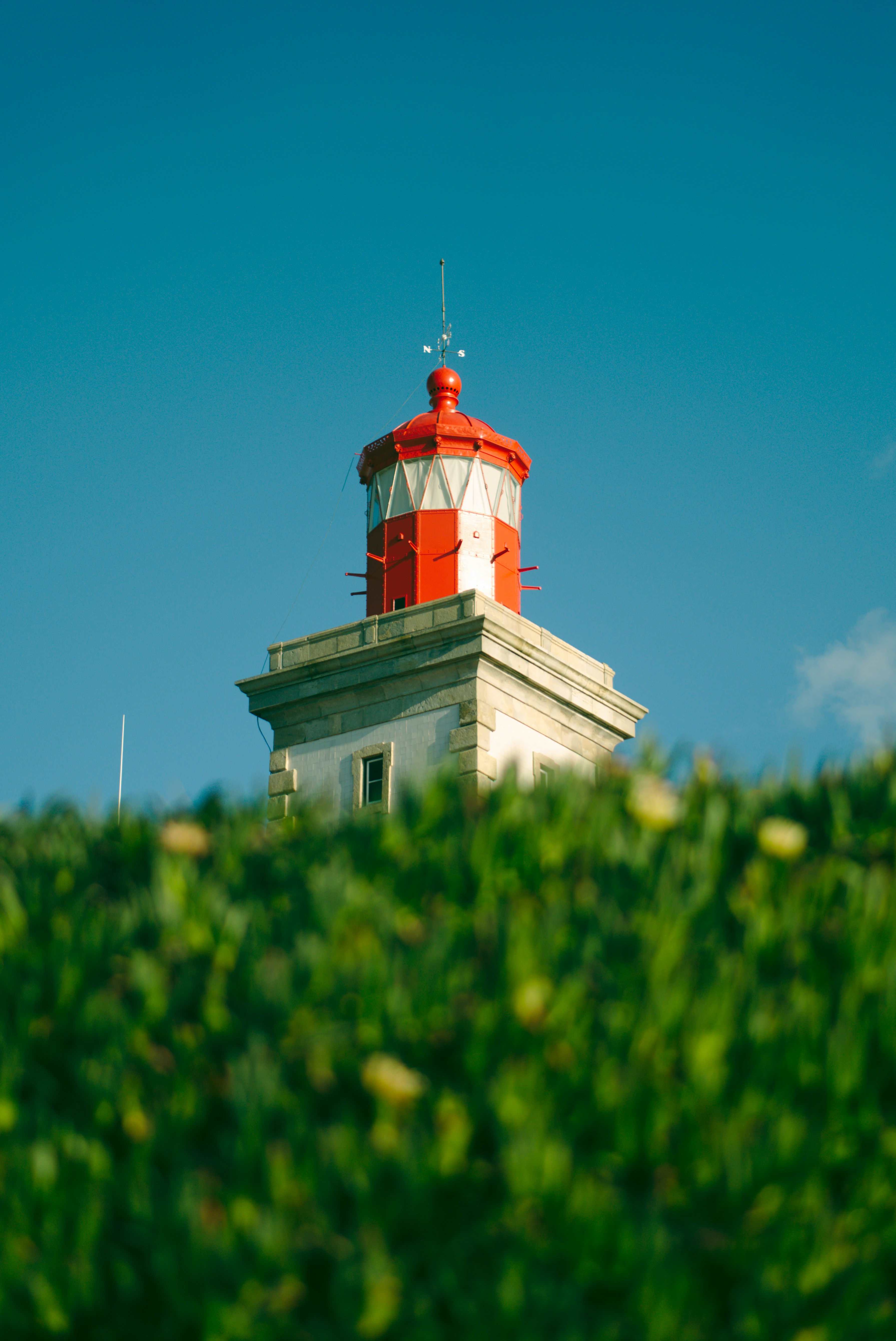 Color photograph of a red-and-white lighthouse perched on a stone building, with a blurred green foreground and a bright blue sky.