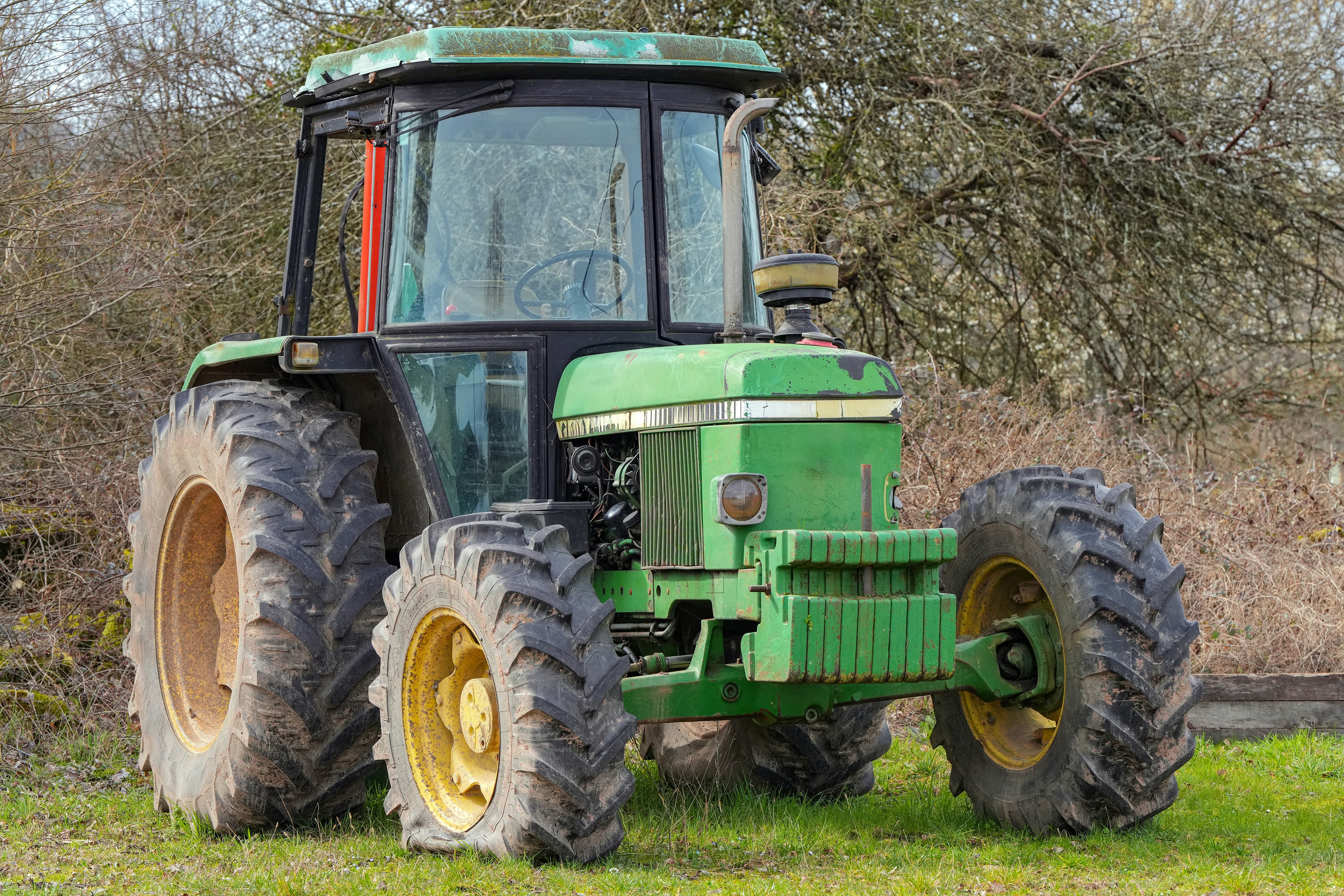 A classic green tractor stands on a grassy field with a backdrop of leafless trees.
