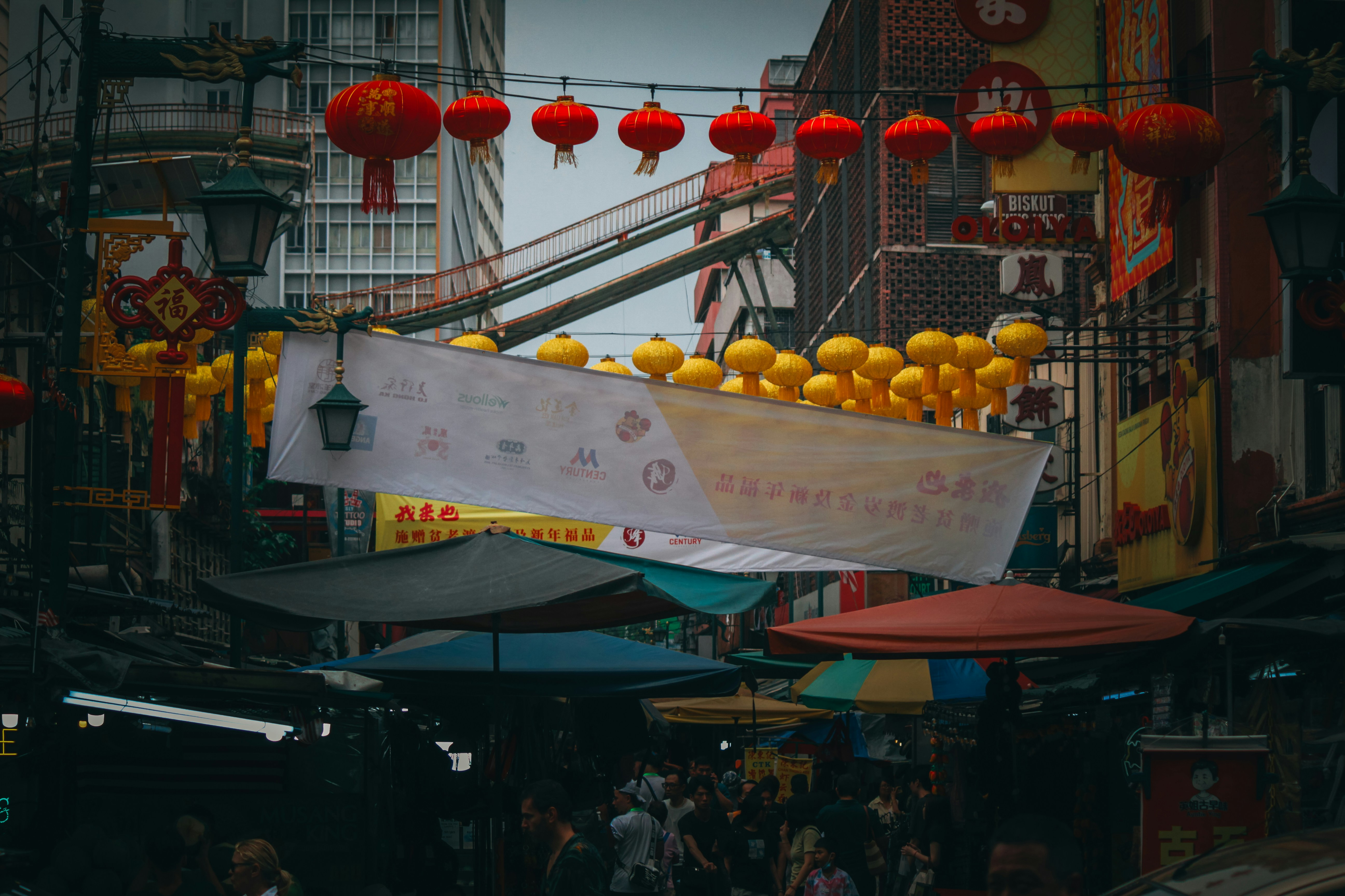 Colorful lanterns hang above a bustling street market, teeming with people and vibrant stalls.