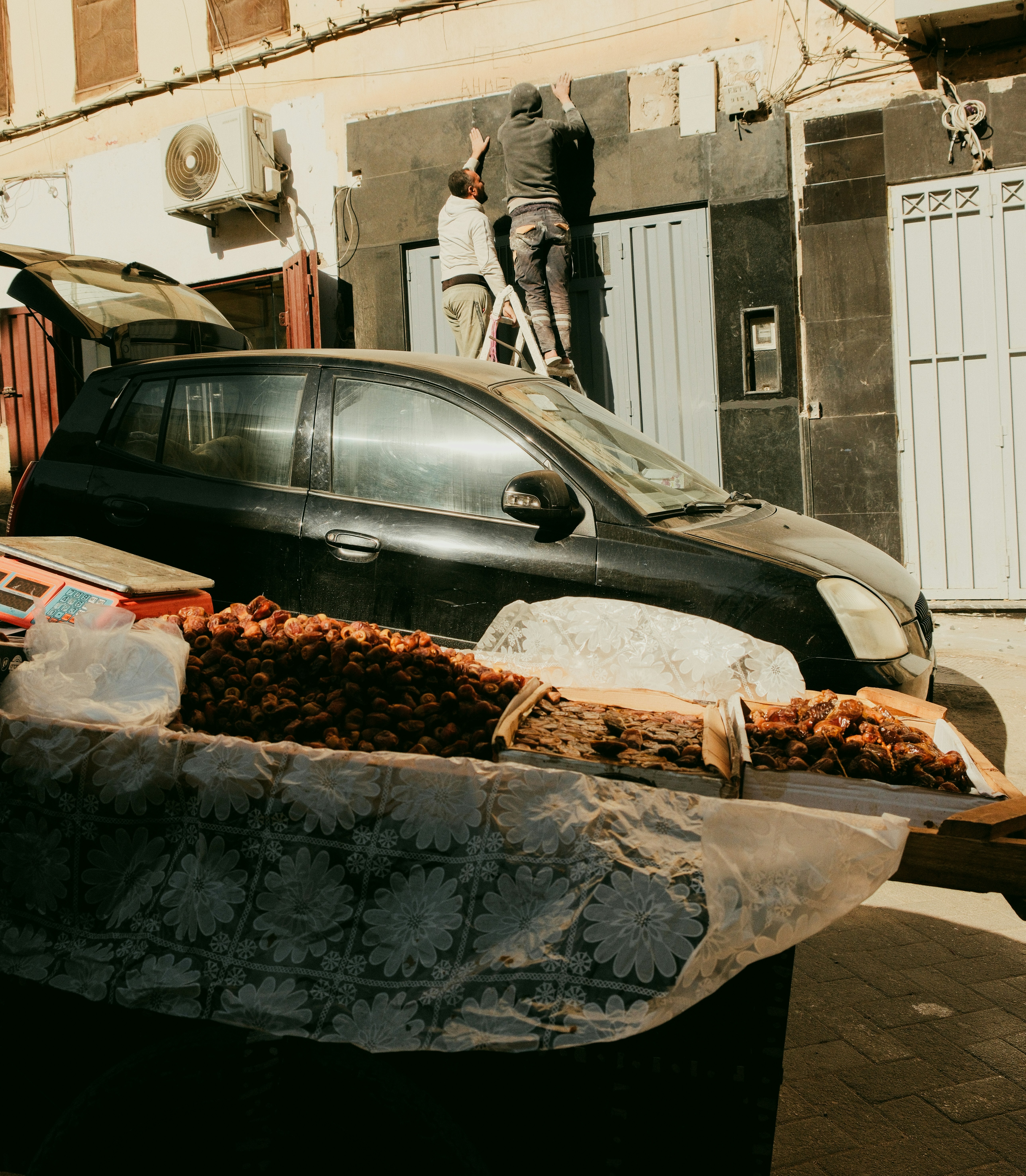Two workers stand on a ladder against a tiled wall beside a parked car, while a vendor’s table piled with dates dominates the foreground.
