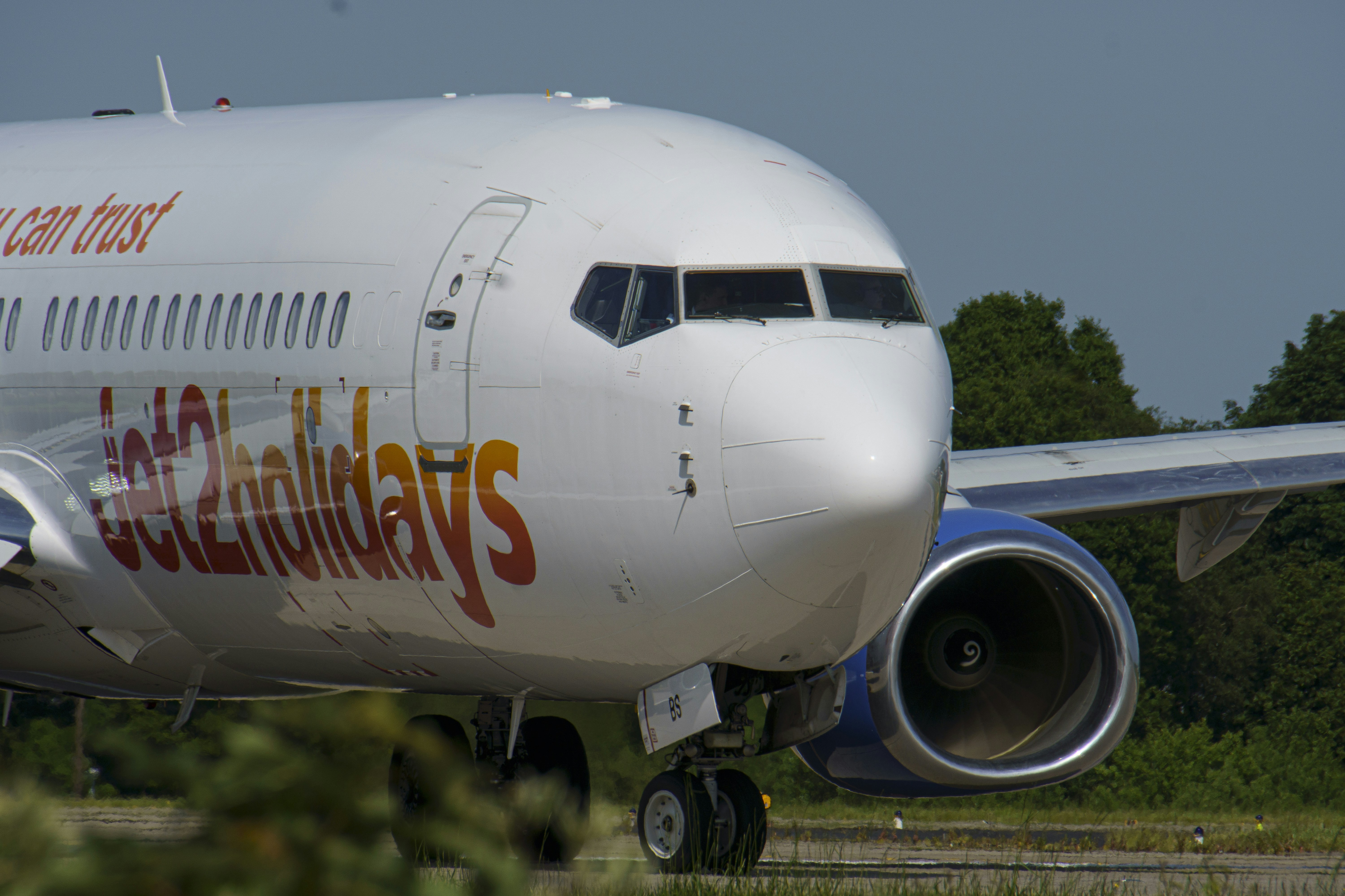 Commercial airplane taxiing on runway with white and blue livery against a clear sky and green trees.