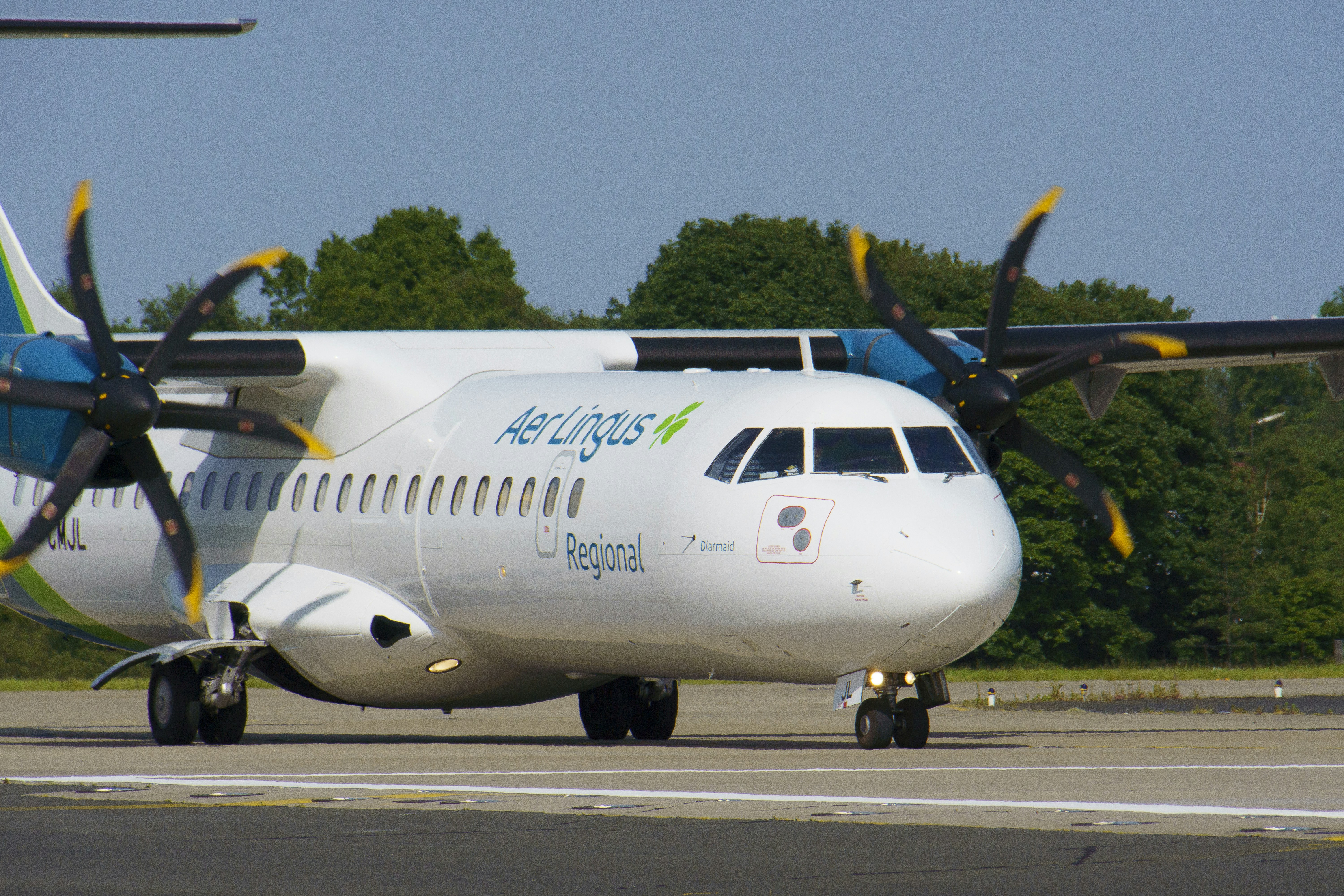 Aer Lingus Regional propeller plane taxiing with a backdrop of green trees and blue sky.