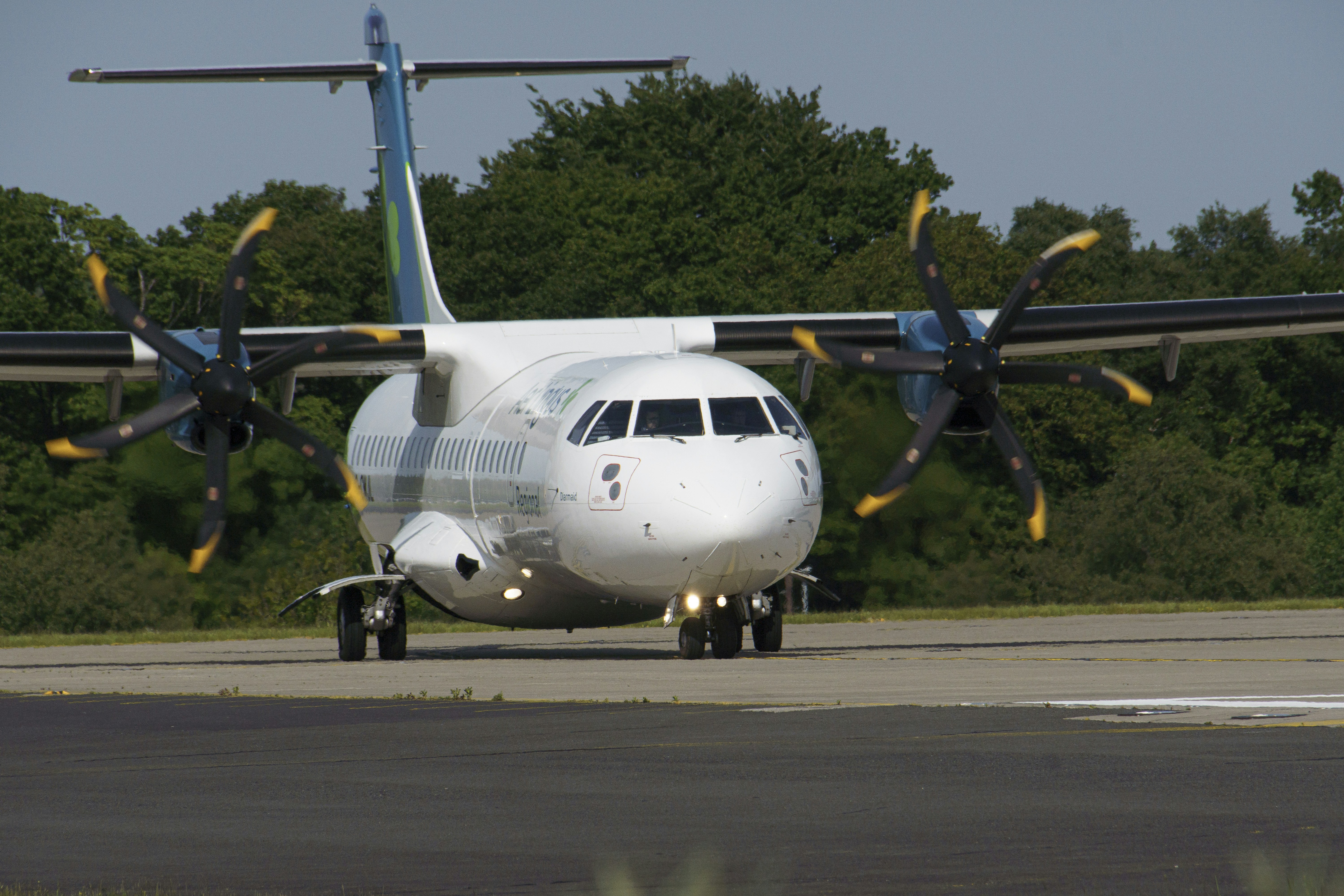 Turboprop plane approaching runway with spinning propellers and green trees in the background.