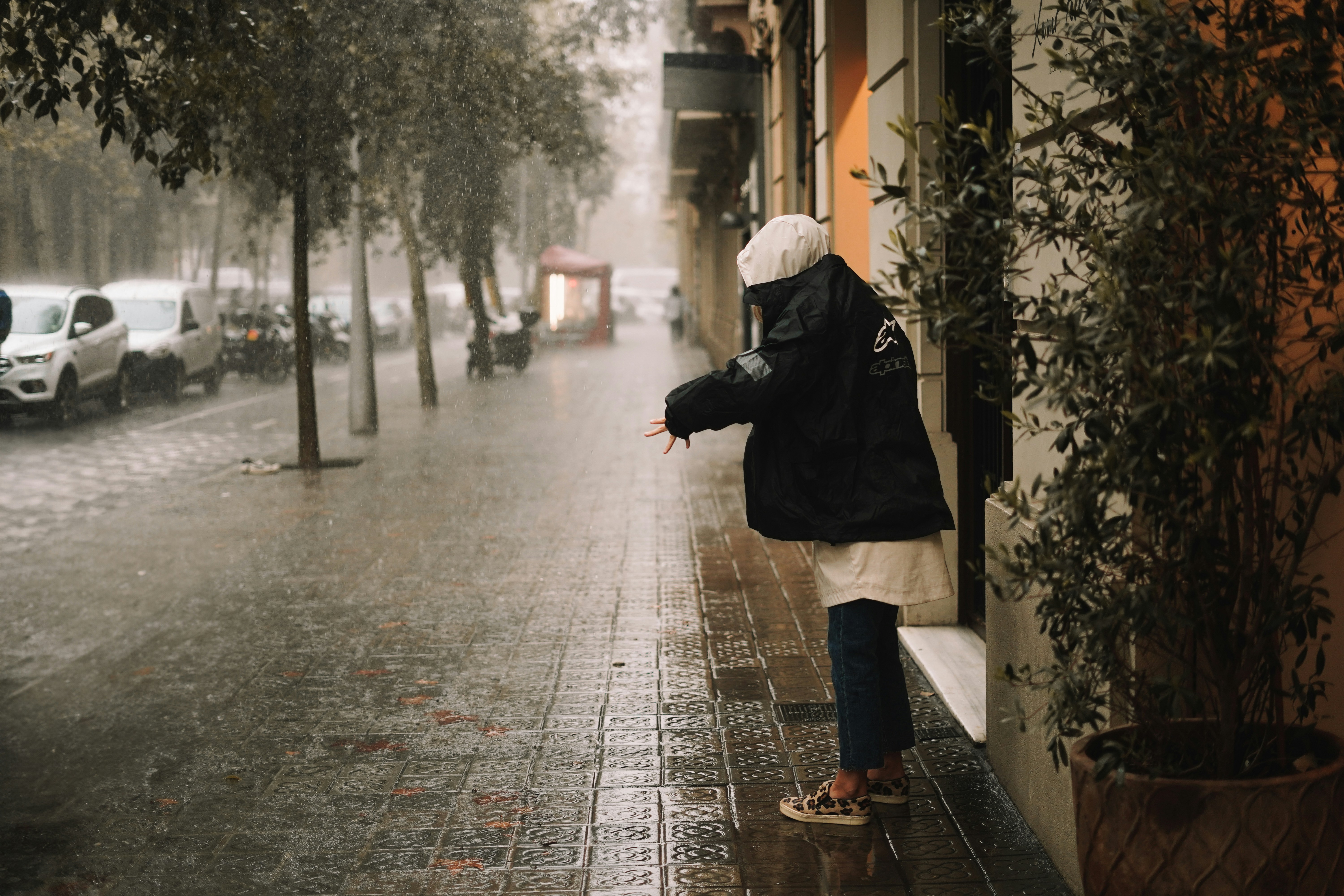 Person in a raincoat reaching out on a wet city sidewalk as rain falls gently.