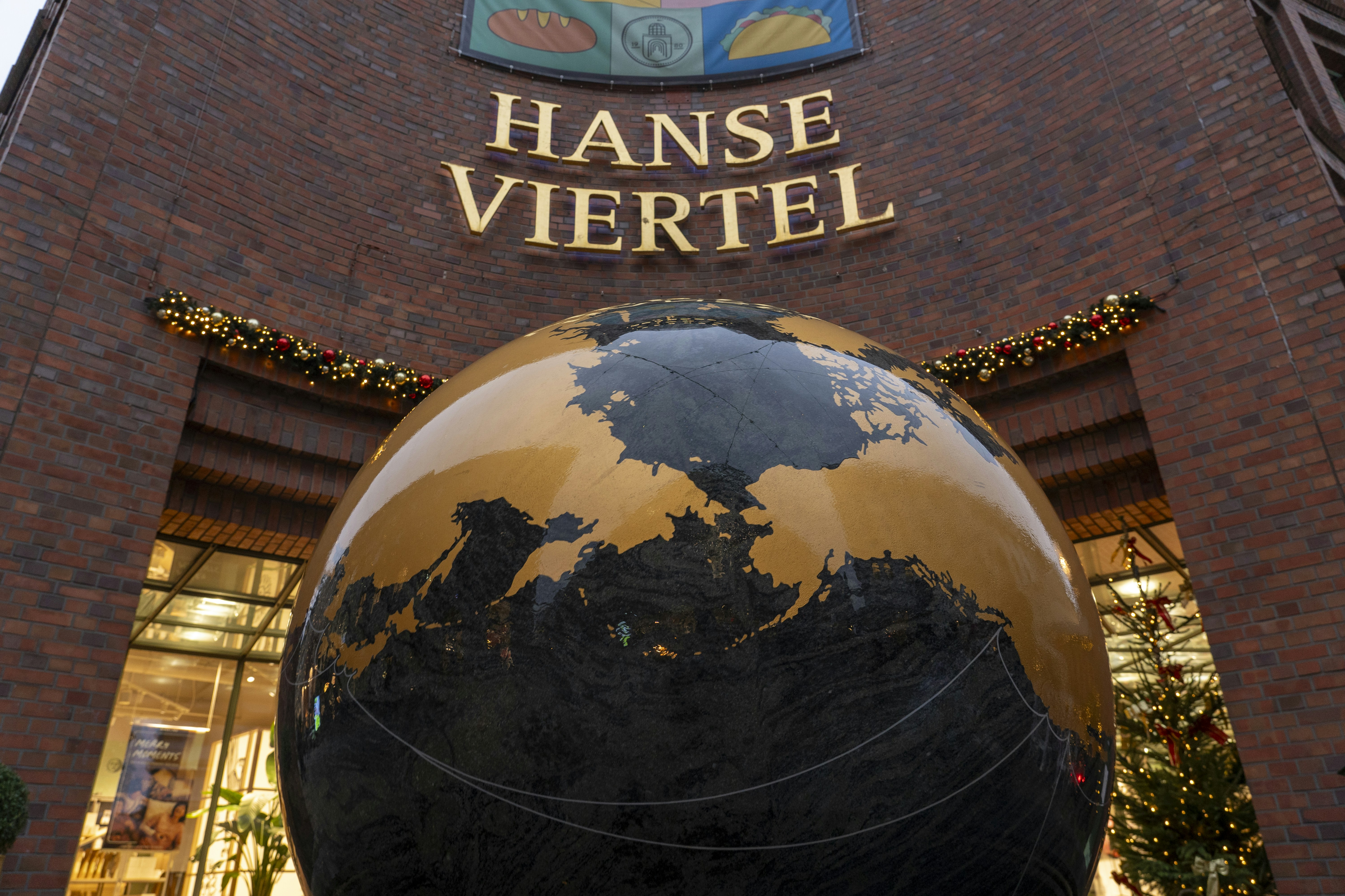 Golden Globe at Hanse Viertel, Hamburg A stunning golden globe sculpture in front of the Hanse Viertel shopping center in Hamburg, Germany. The brick facade adorned with festive Christmas decorations creates a warm and inviting holiday ambiance, highlighting the vibrant city atmosphere.