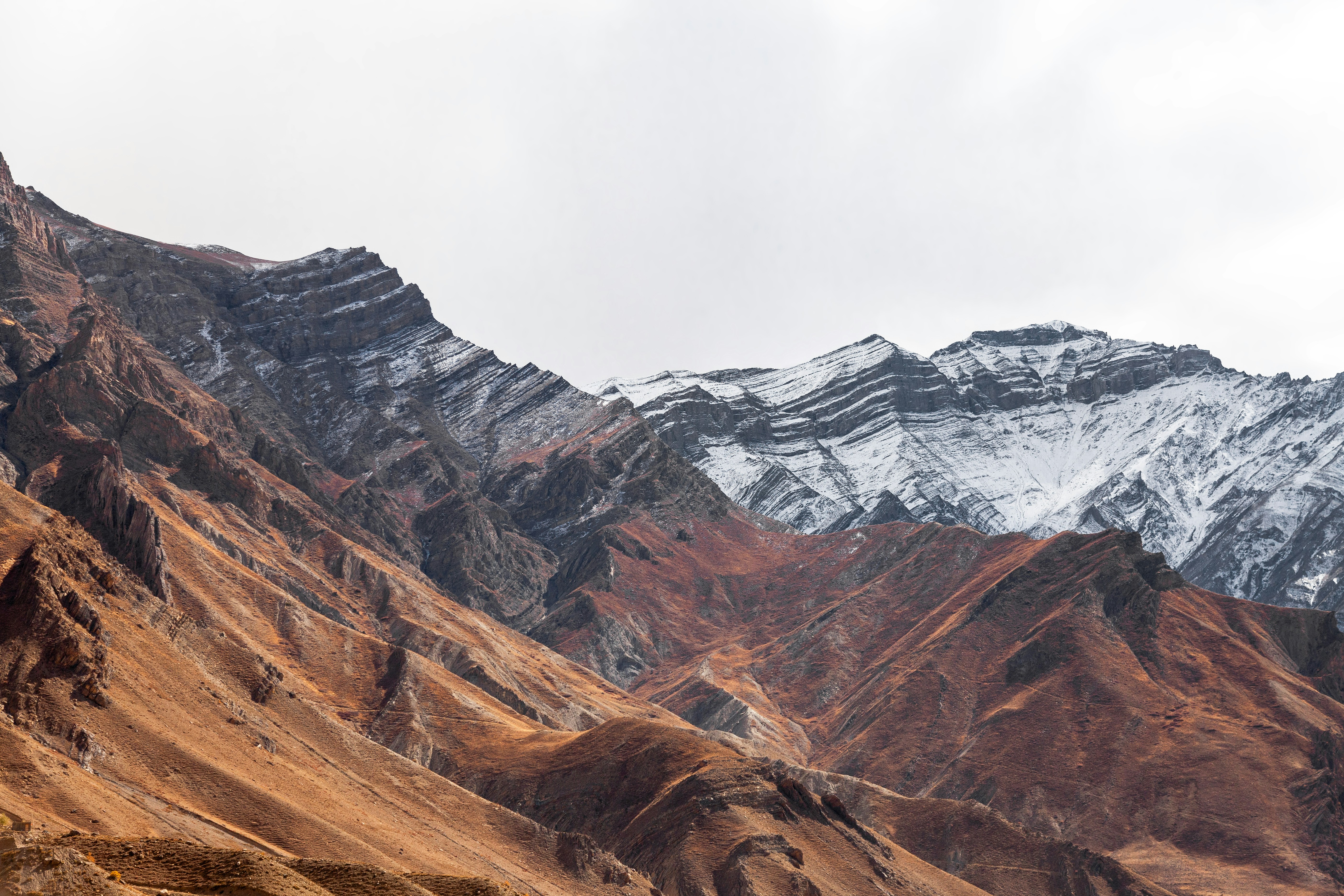 Layered mountain range with snow-dusted peaks and reddish-brown slopes under an overcast sky.