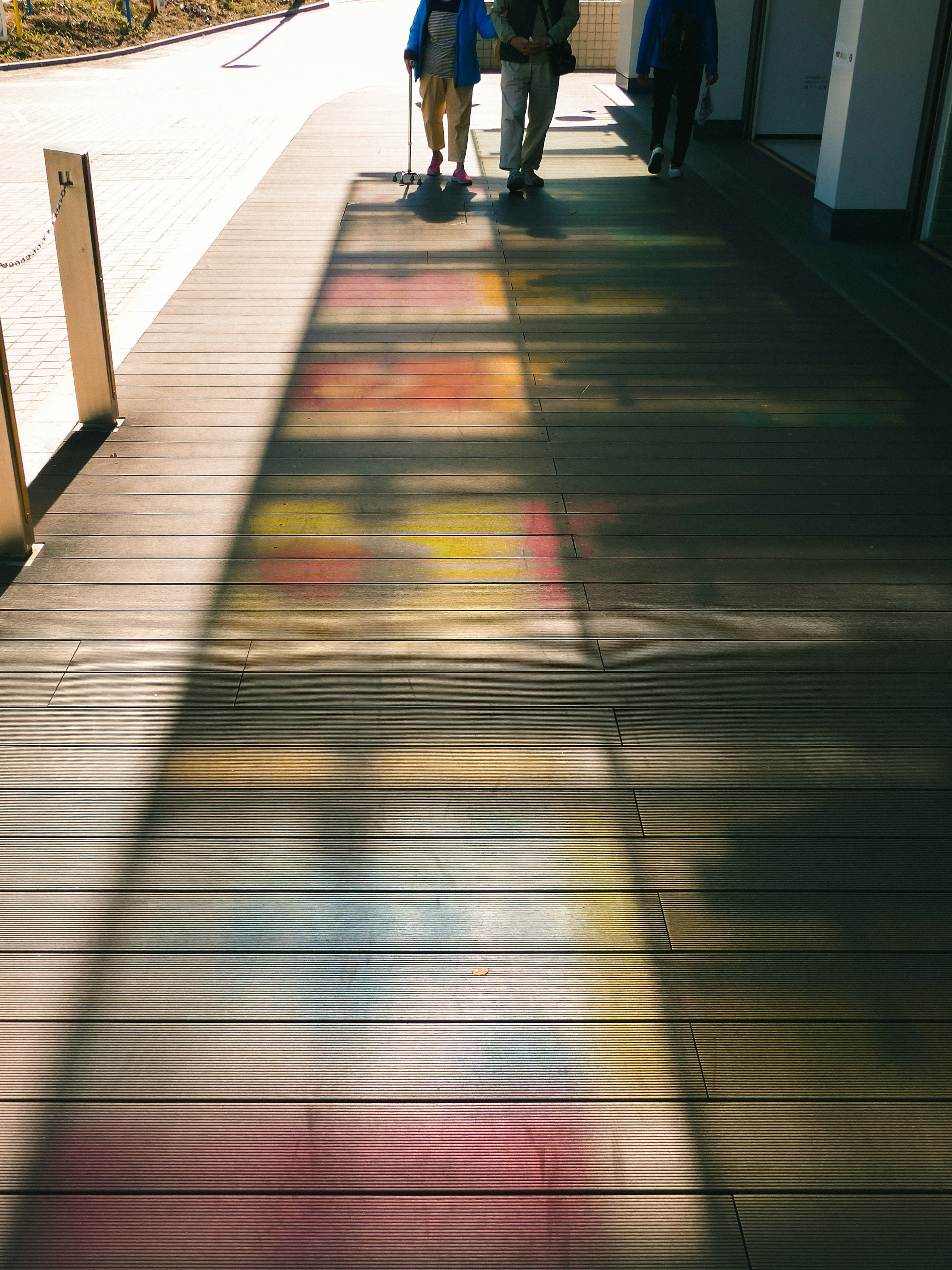 Sunlit wooden boardwalk shows rainbow-colored reflections on the planks while three people walk toward a building, their shadows stretching diagonally across the scene.