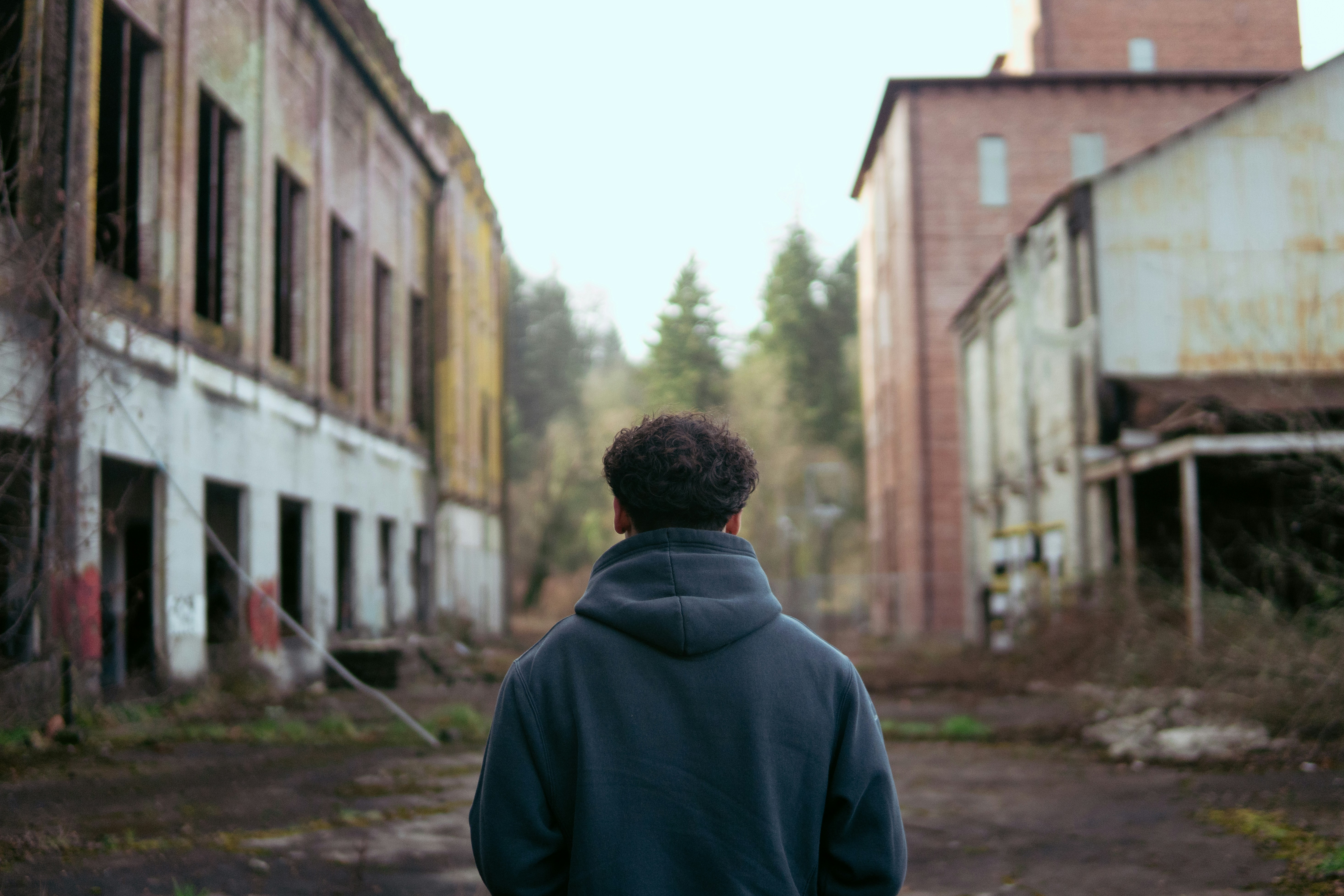 Person facing weathered industrial buildings with overgrown greenery in the background.