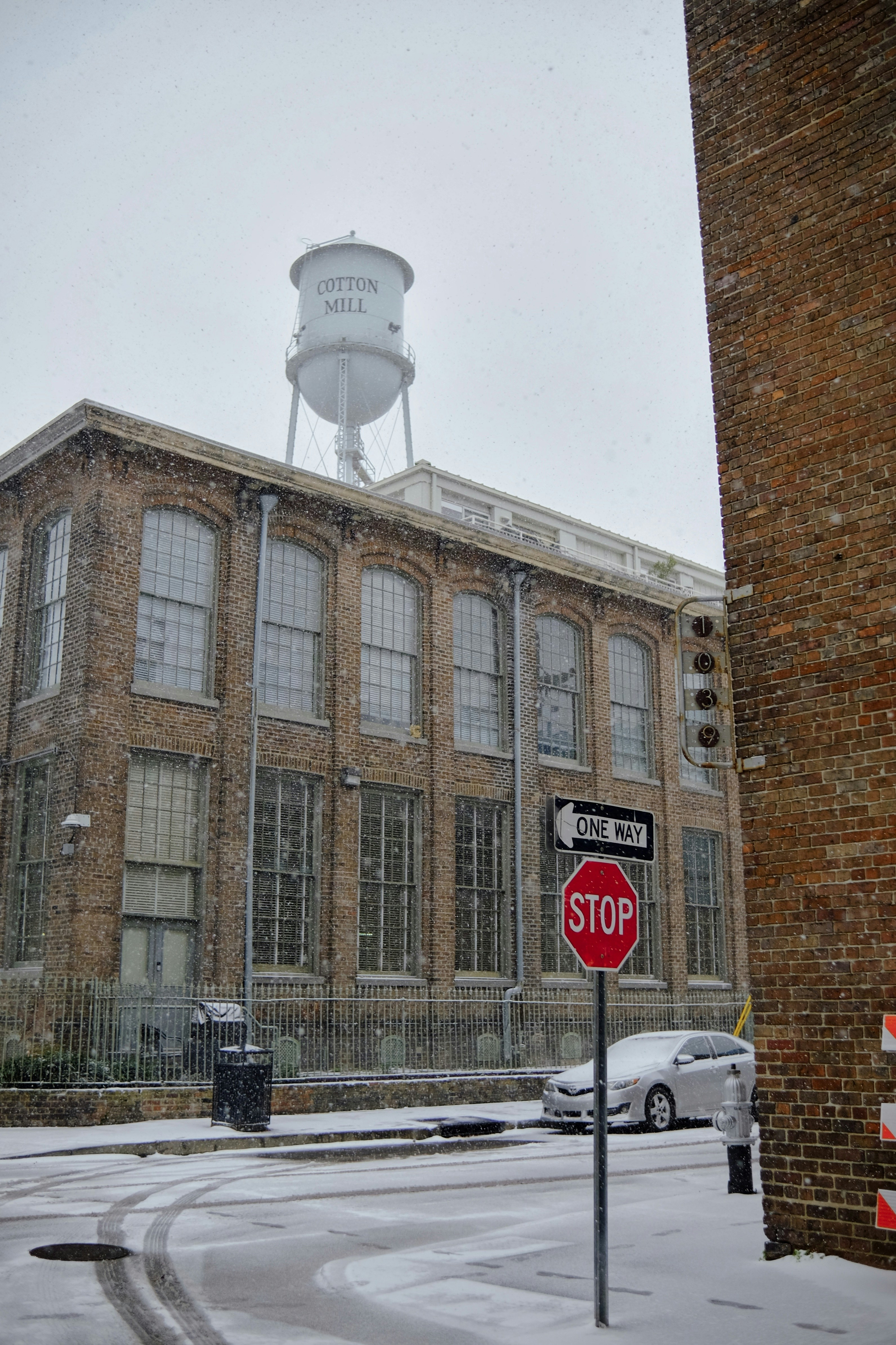 Snow blankets a brick Cotton Mill building as a tall water tower rises above; a stop sign and a parked silver car anchor the quiet urban scene.