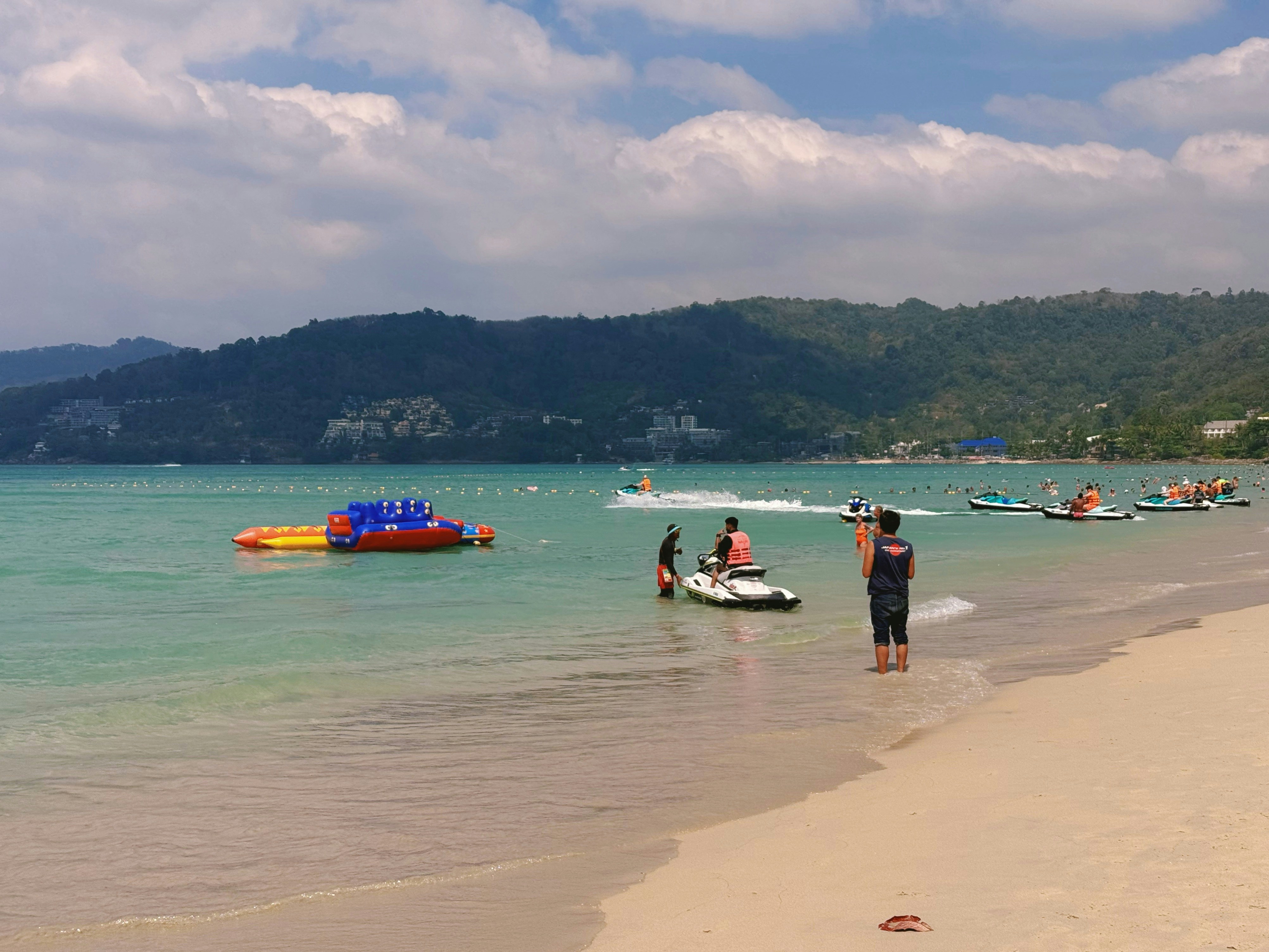 Beachgoers enjoy jet skis and inflatable rides on the turquoise waters of Patong Beach under a partly cloudy sky.