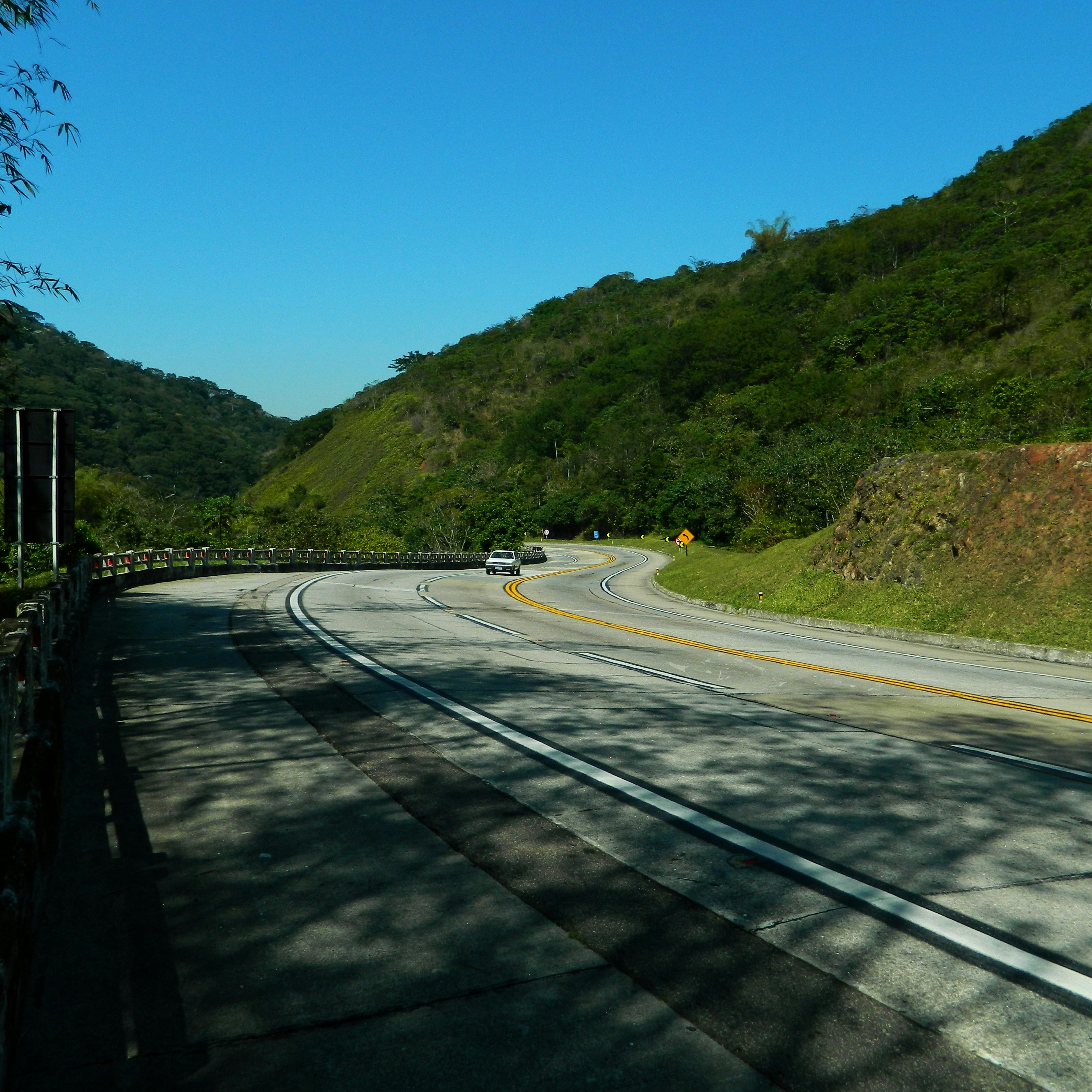 Sunlit mountain road curves through green hills with guardrails on the left and a distant white car approaching the bend.