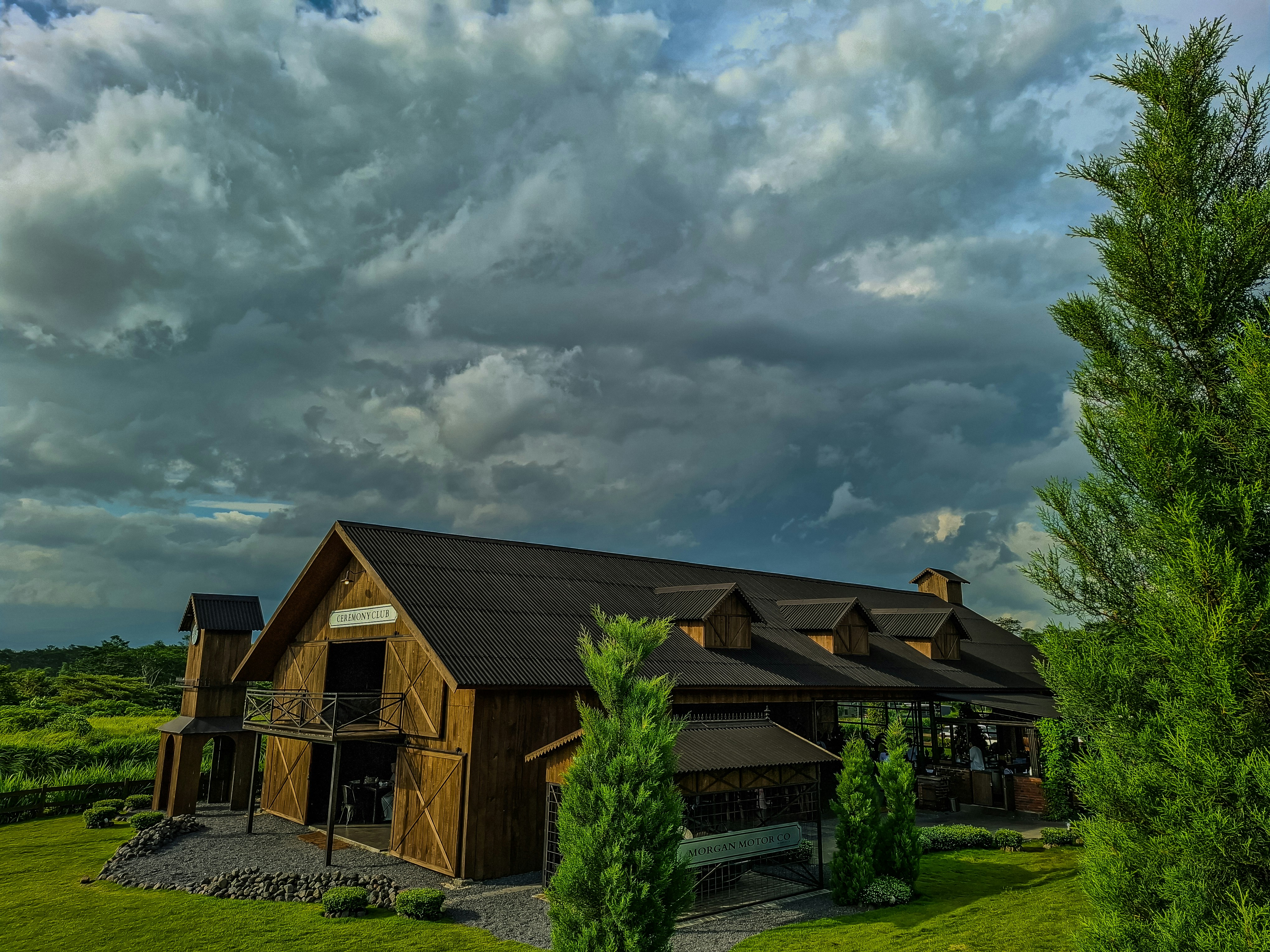 Rustic wooden lodge sits amid a grassy field beneath dramatic, cloud-filled skies, with evergreen trees framing the scene.