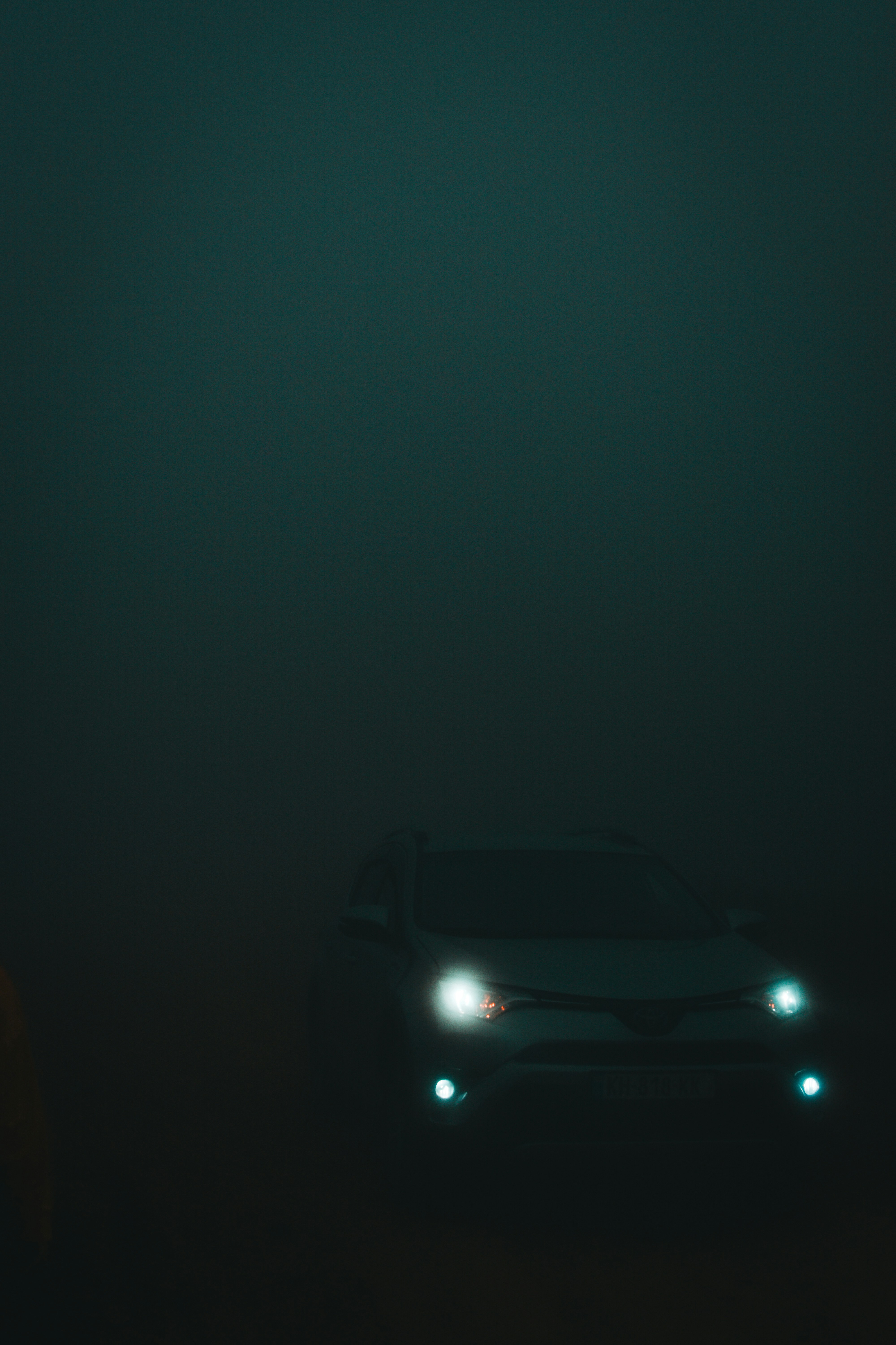 A lone car's headlights pierce through dense fog, creating an eerie atmosphere that envelops the scene. The obscured surroundings enhance the sense of isolation.
