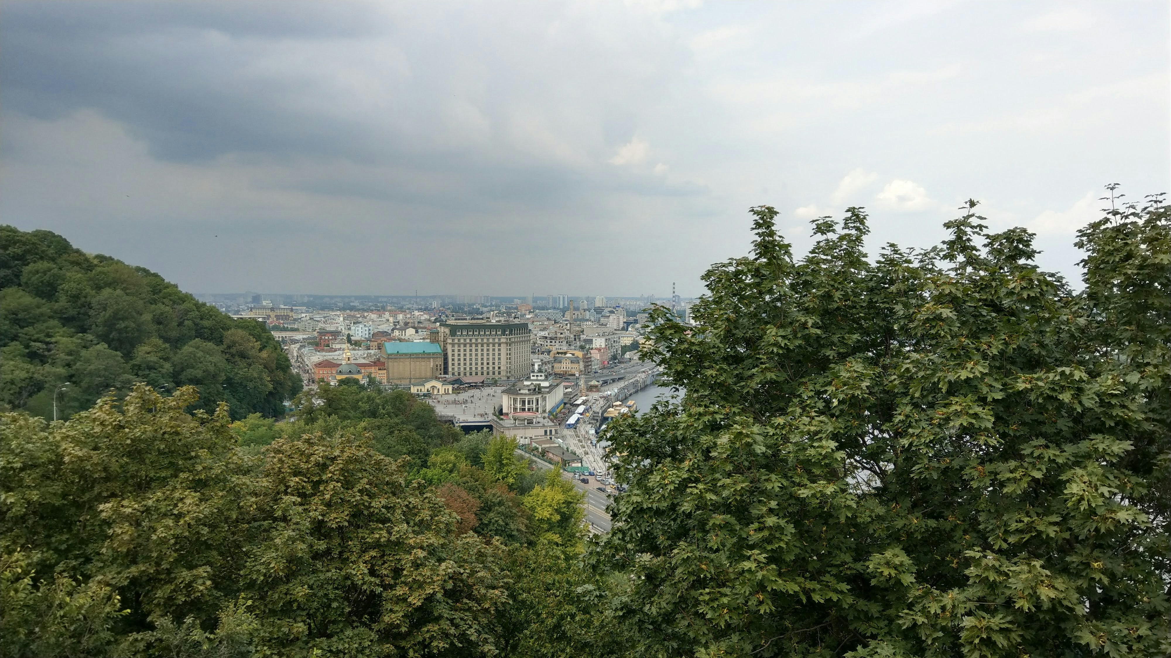 Cityscape view from a hilltop framed by lush green trees under a cloudy sky.