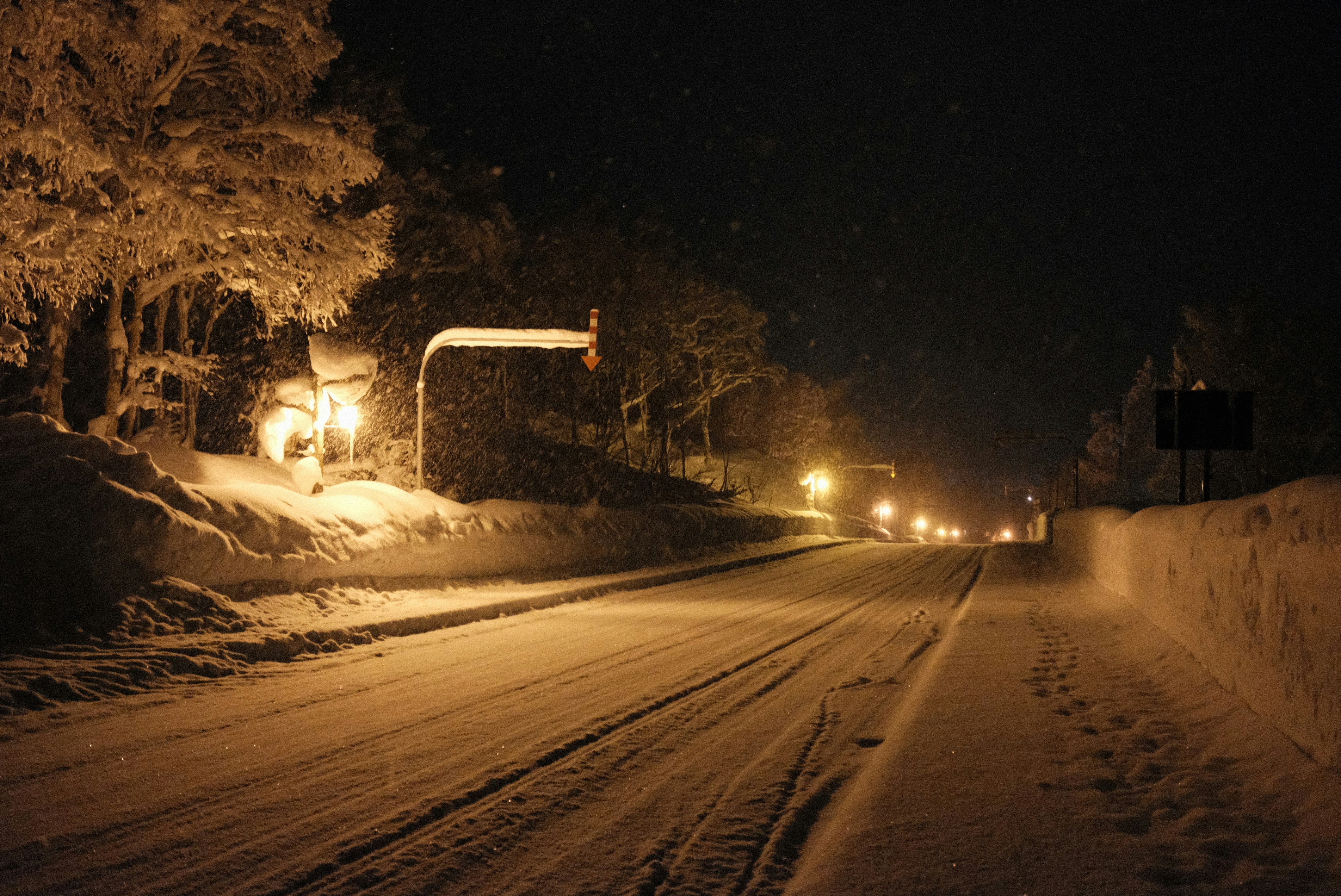 Snow-covered road lit by warm streetlights, creating a serene winter night scene with trees and a distant glow.