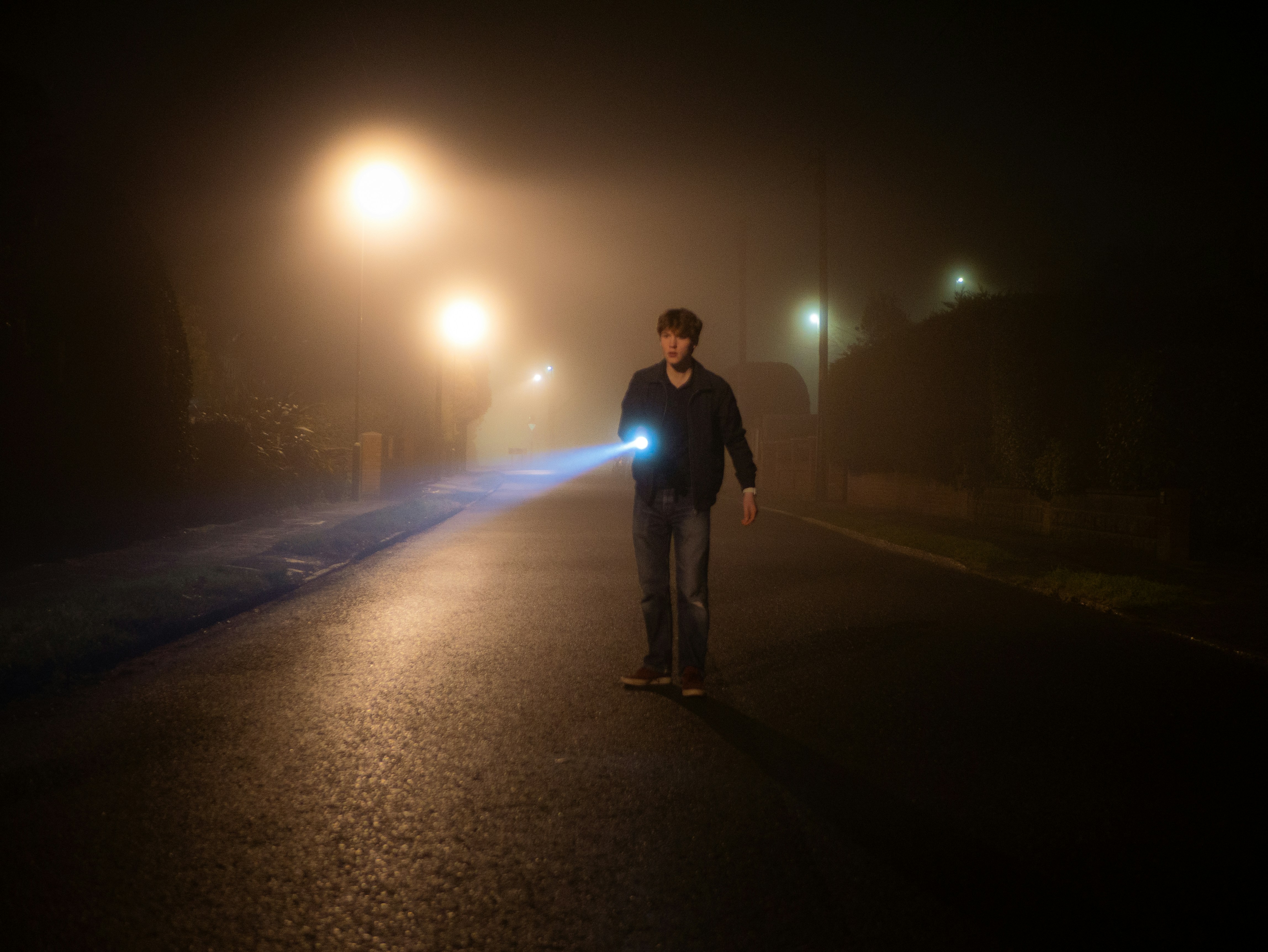 A lone figure navigates a misty street with a flashlight, while streetlights cast an ethereal glow through the fog.